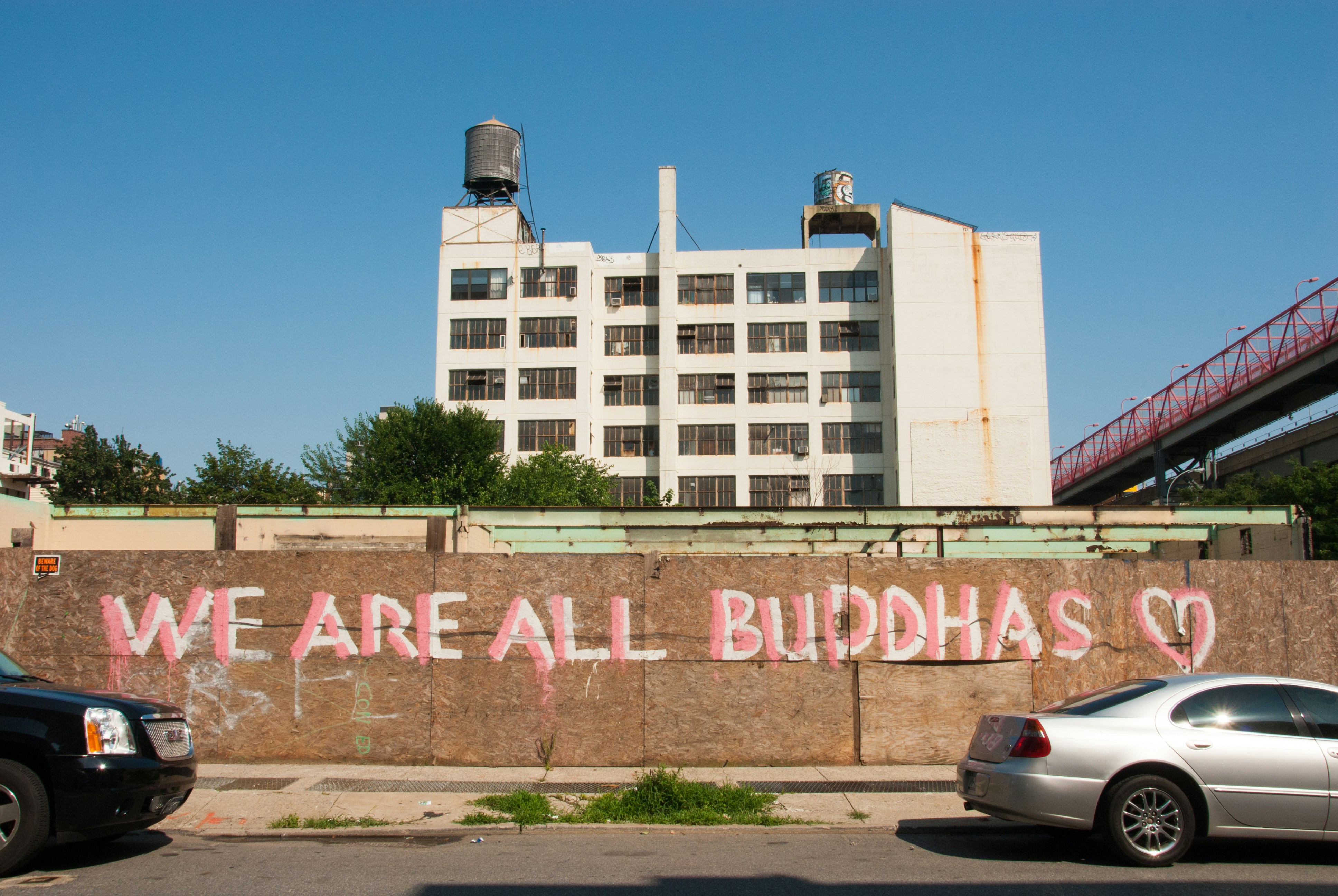 Graffiti reading 'We Are All Buddhas' on a wall below a white concrete building under a clear blue sky.