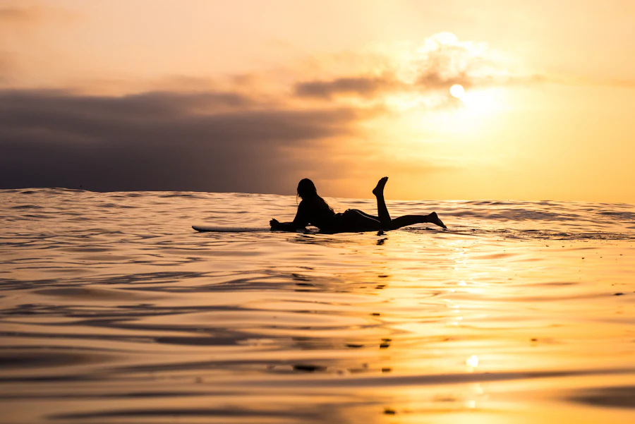 Surfer lying on a board, silhouetted against a vibrant sunset over calm waters. The scene captures the tranquil essence of evening surf.