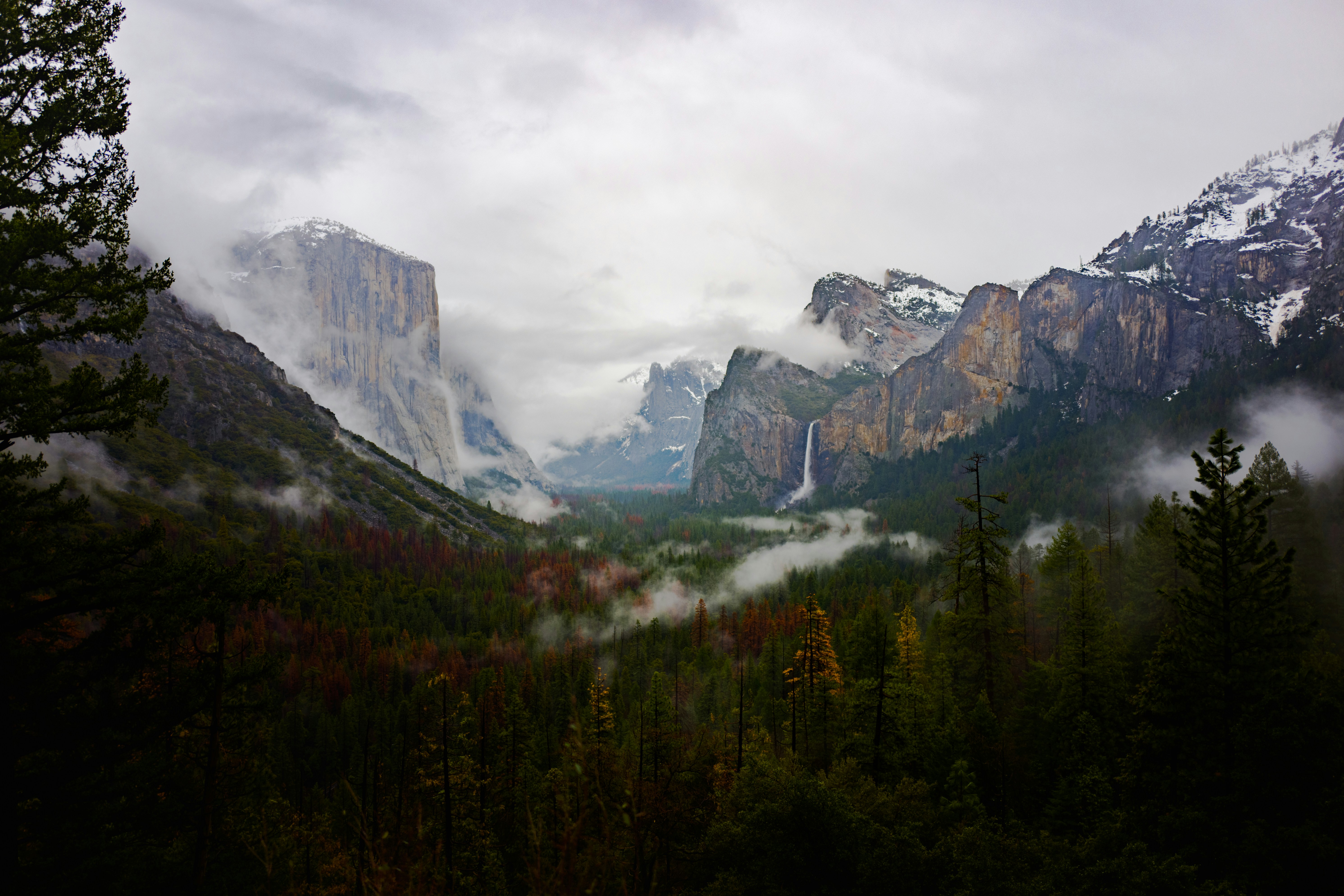 Dramatic mountain landscape shrouded in mist, featuring towering cliffs and a cascading waterfall amidst a lush forest. The scene evokes a sense of tranquility and awe.