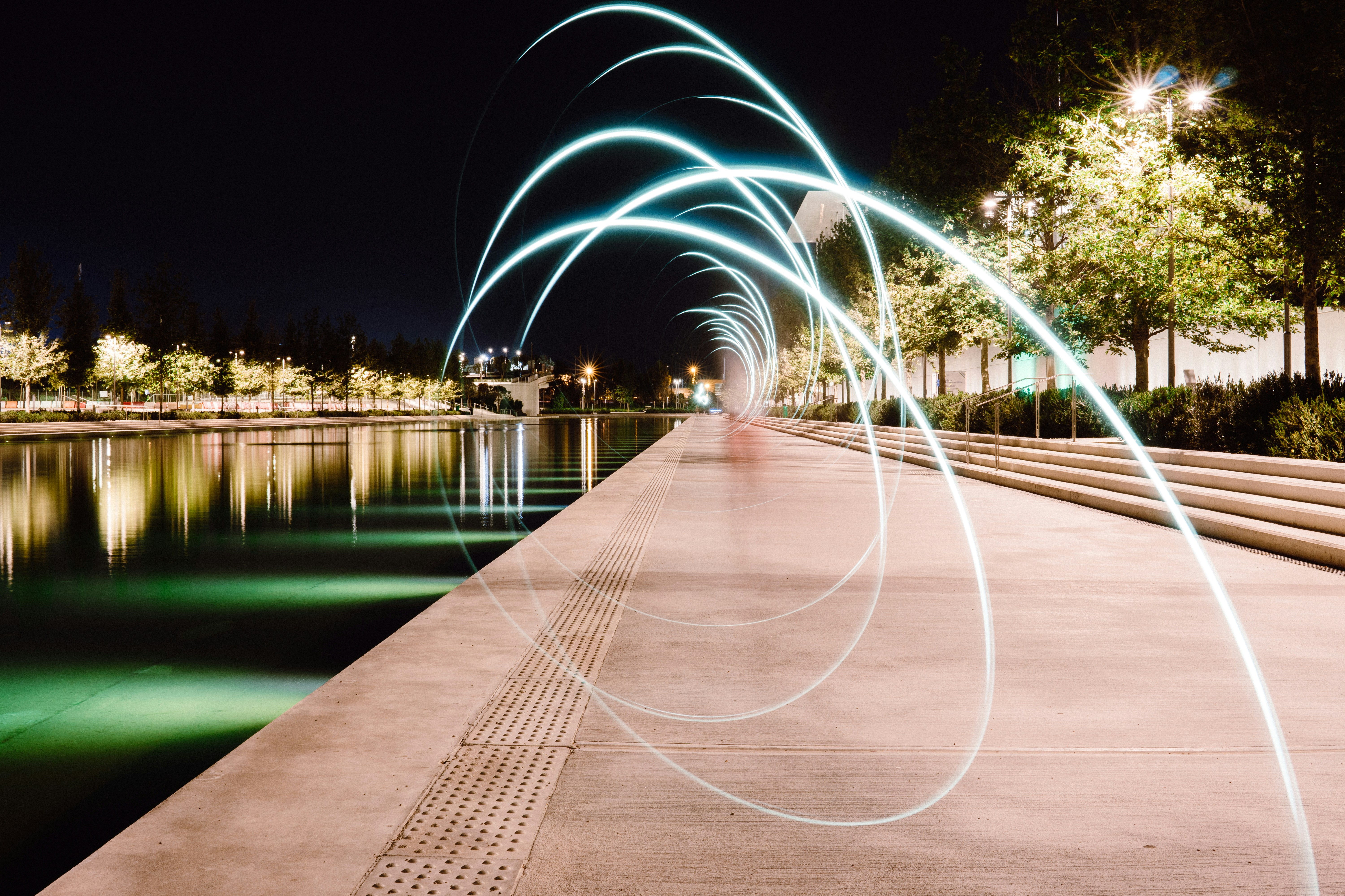 Long exposure of light trails along a riverside promenade at night, with trees and water reflecting ambient light.