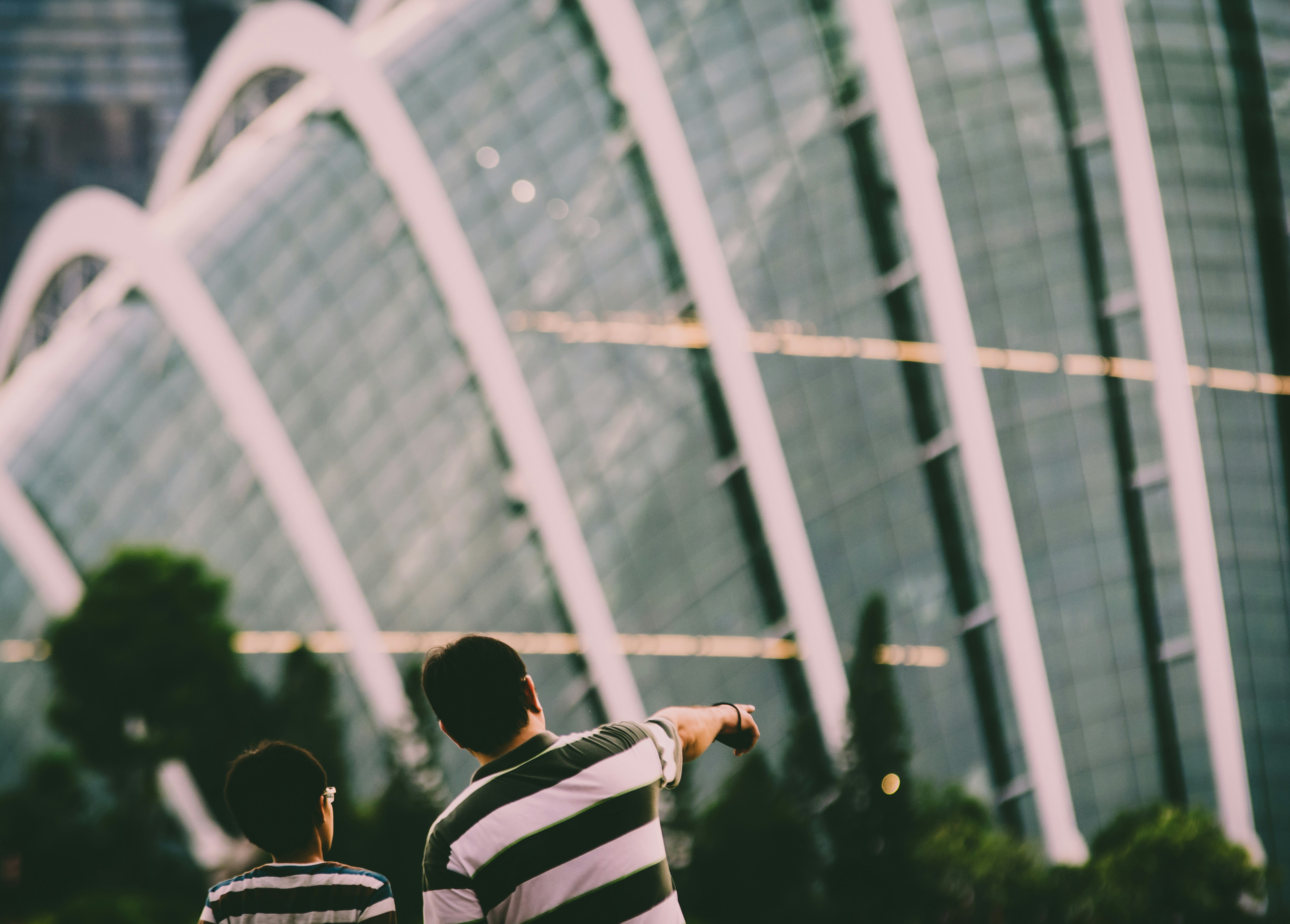 Two individuals in striped shirts admire a modern glass structure, pointing towards its intricate design. The scene captures a moment of exploration and wonder.