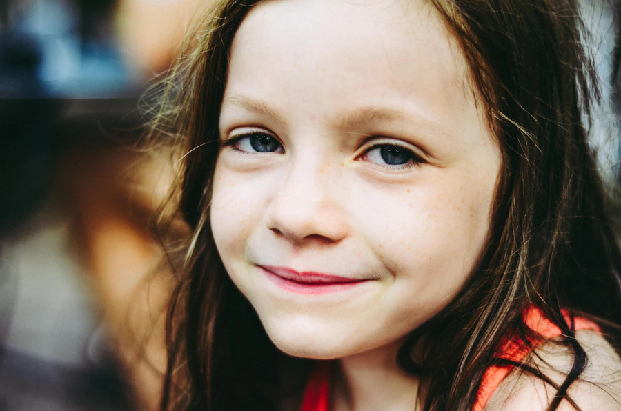 Young girl with long brown hair and a gentle smile, wearing a vibrant red top. The background features soft, blurred colors, enhancing her cheerful expression.