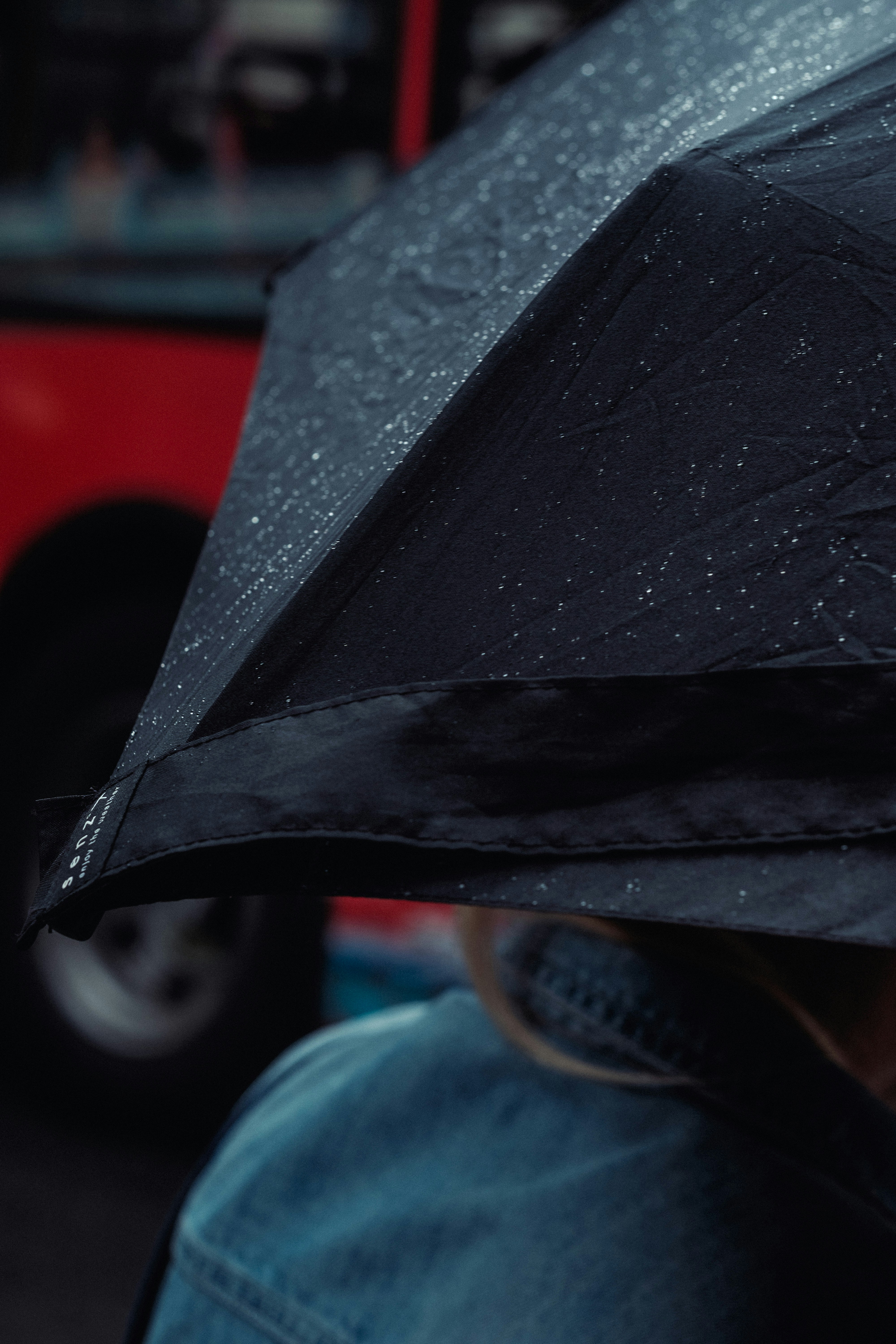 Close-up of a black umbrella speckled with raindrops, with hints of a blurred urban background. The scene conveys a sense of solitude amidst a bustling environment.