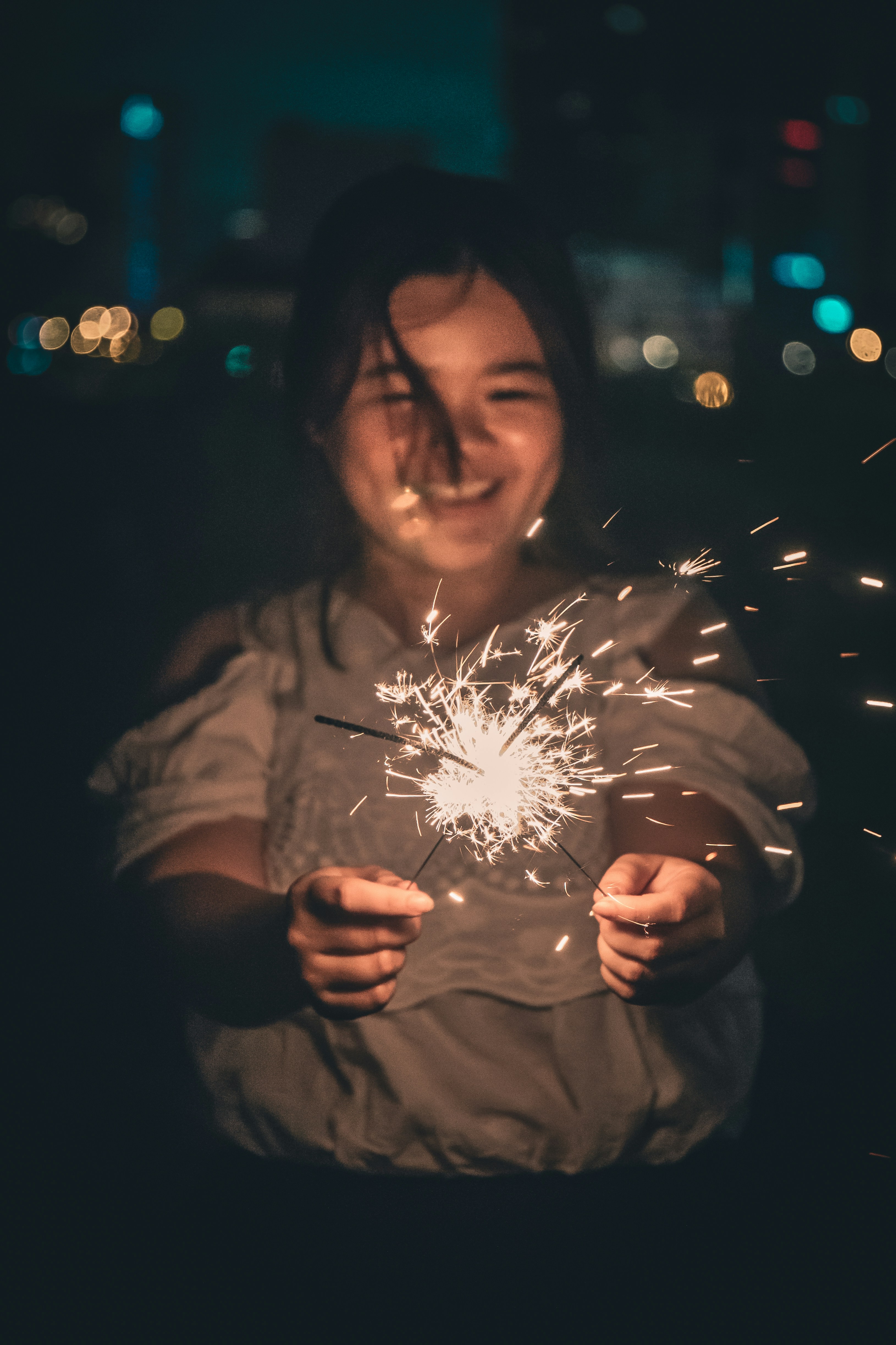 A young woman joyfully holds a sparkler, its bright sparks contrasting against the dark background of a city skyline at night.