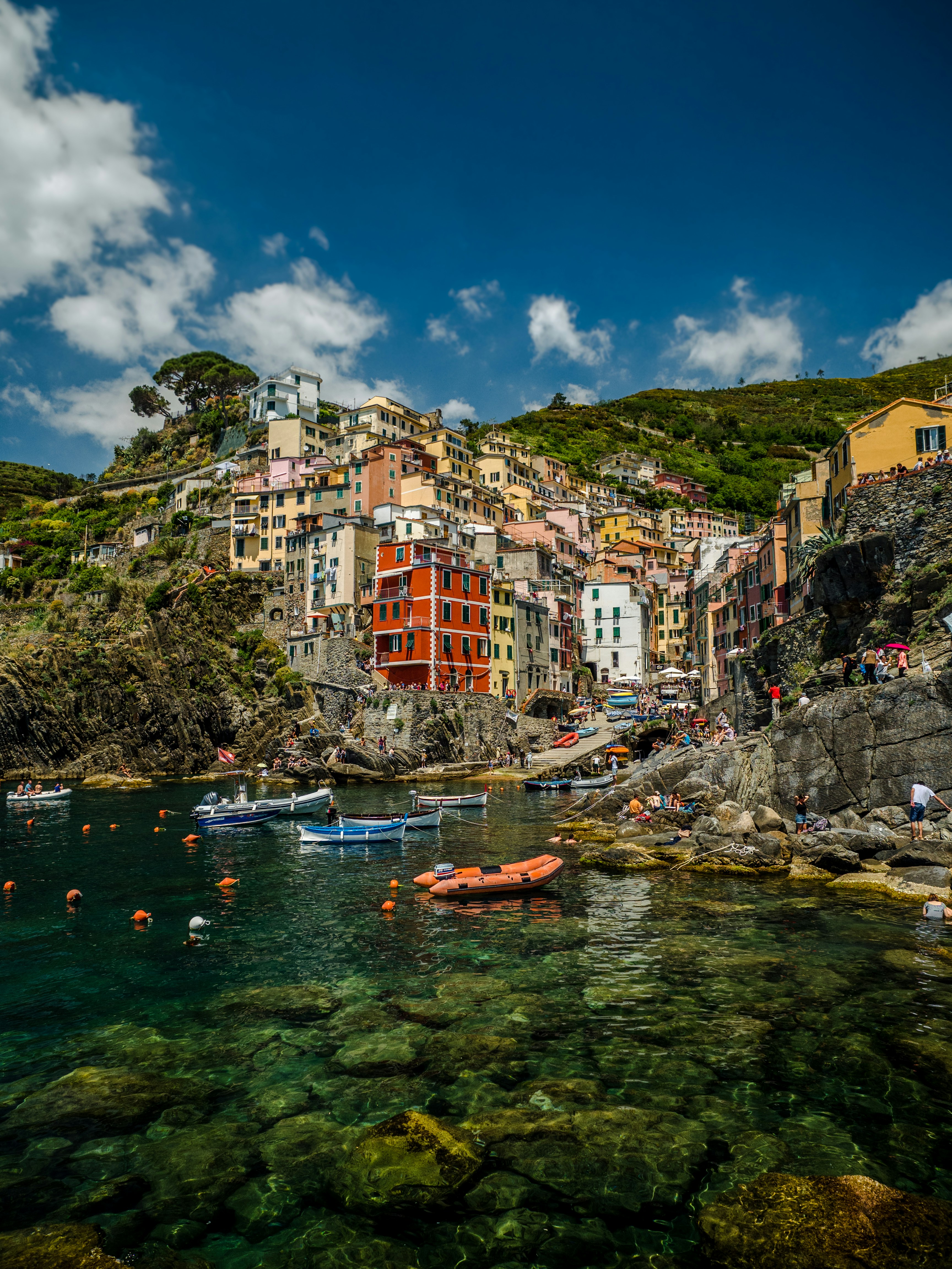 Colorful buildings cascade down a rocky hillside into a tranquil harbor, where boats float among the clear waters and vibrant buoys. The scene captures the charm of a coastal village nestled in nature.