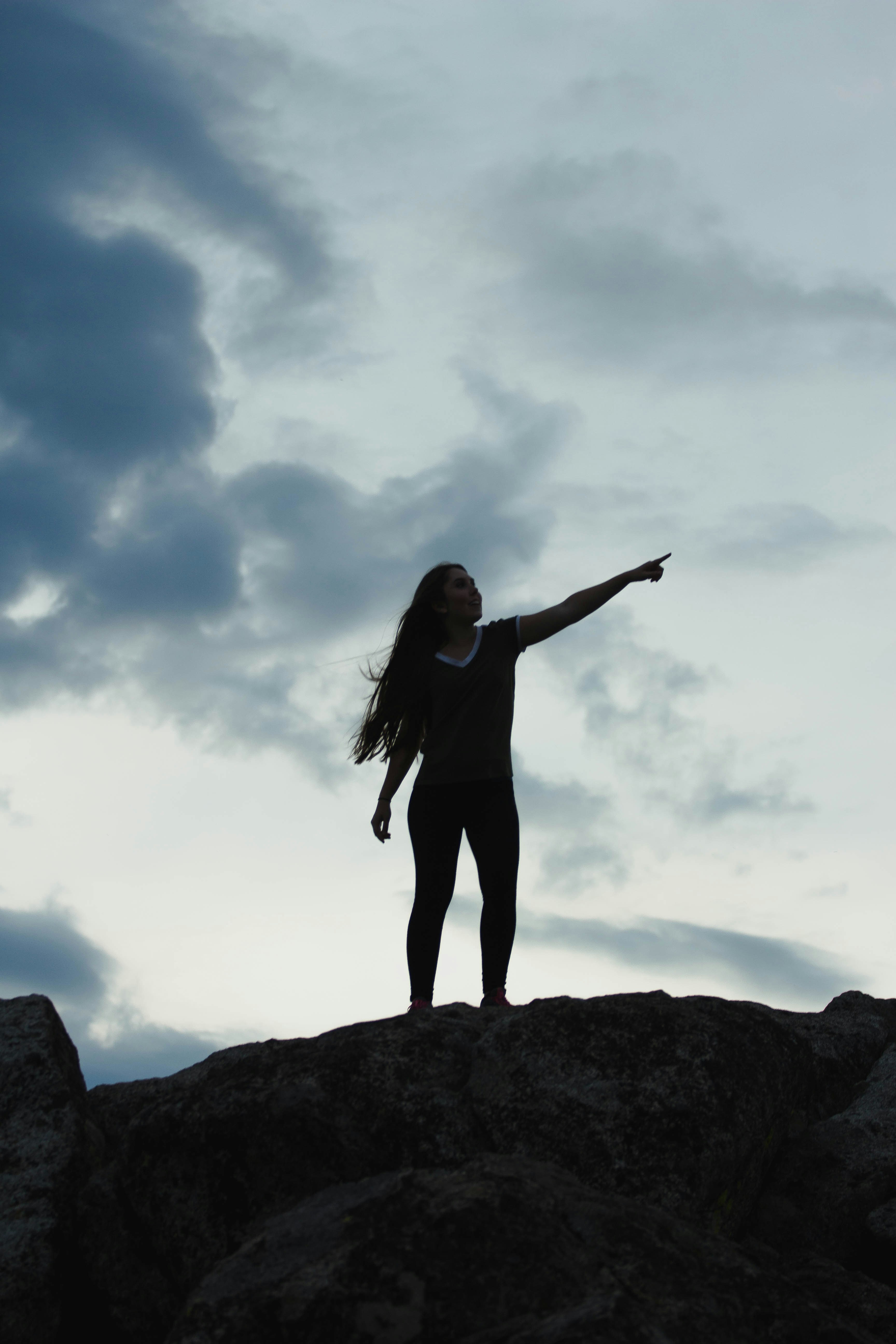 Silhouette of a woman pointing towards the sky while standing on a rocky cliff under a cloudy sky.