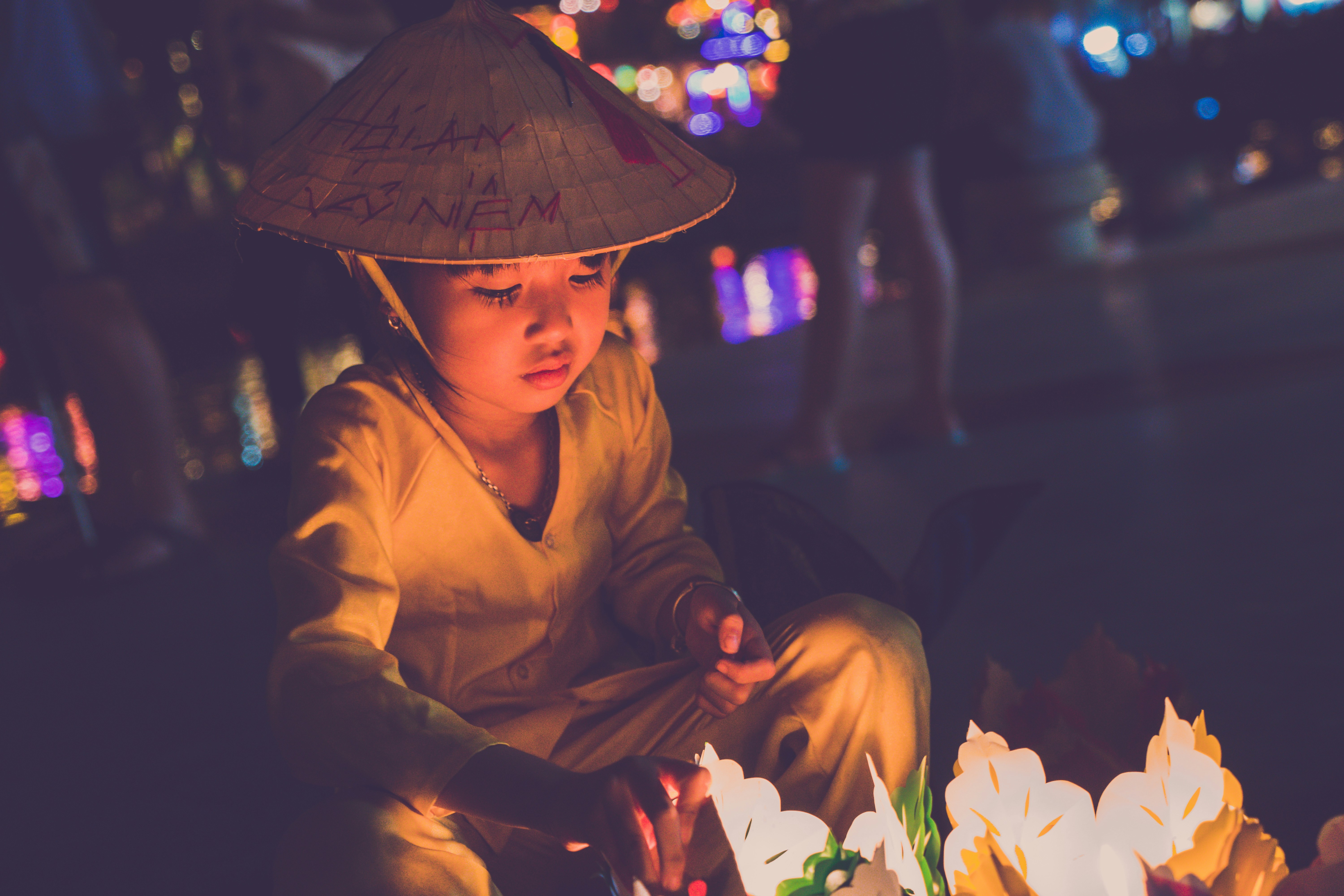 A child in traditional attire delicately arranges glowing lanterns by a serene waterway during a nighttime festival.
