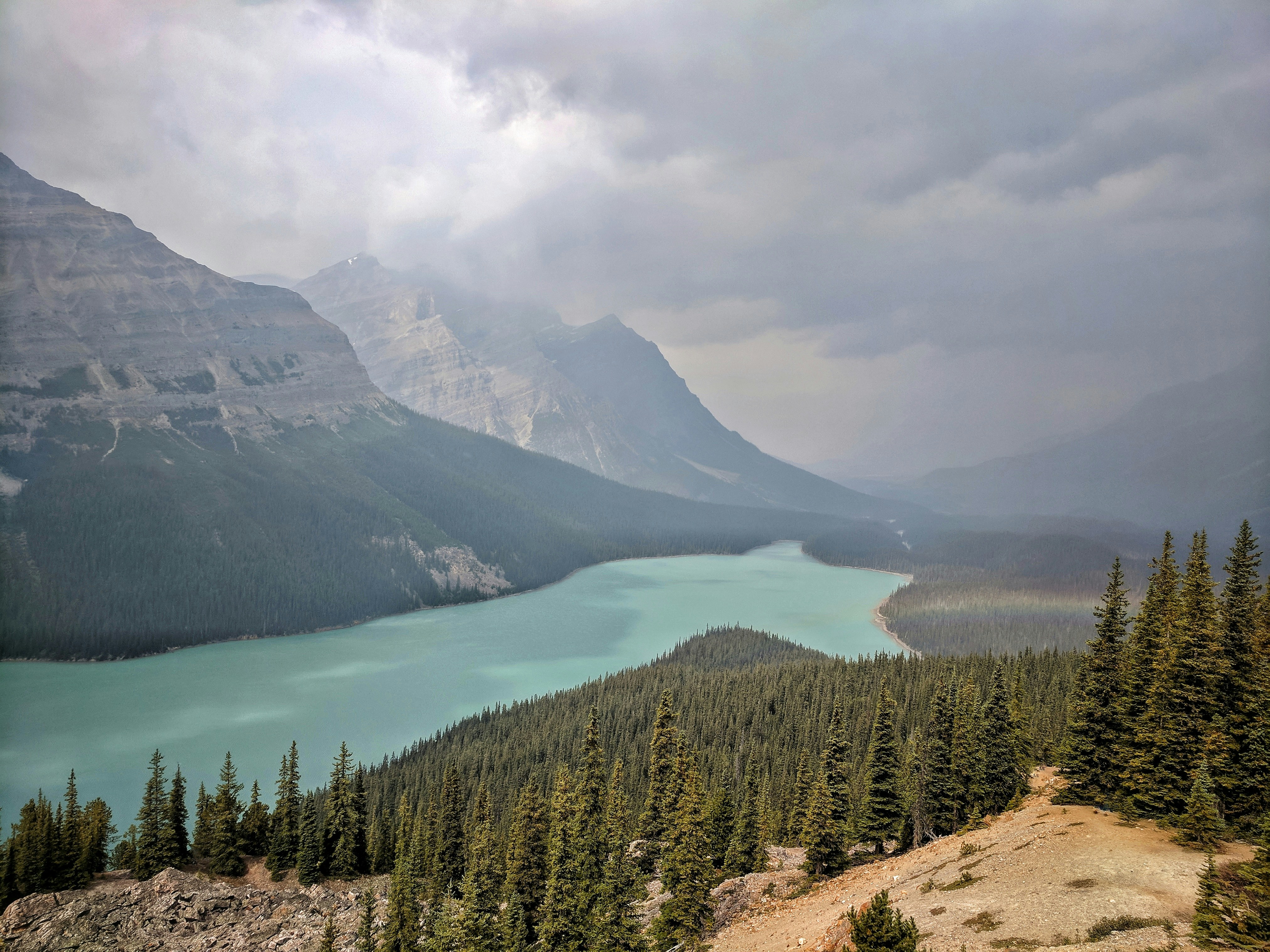 Turquoise lake winding through rugged mountains under a moody sky, framed by evergreen forests. 