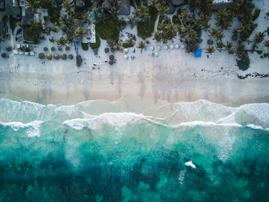 Aerial view of waves breaking onto a sandy tropical beach bordered by palm trees and scattered umbrellas. Clear turquoise water fades into deeper blue offshore.