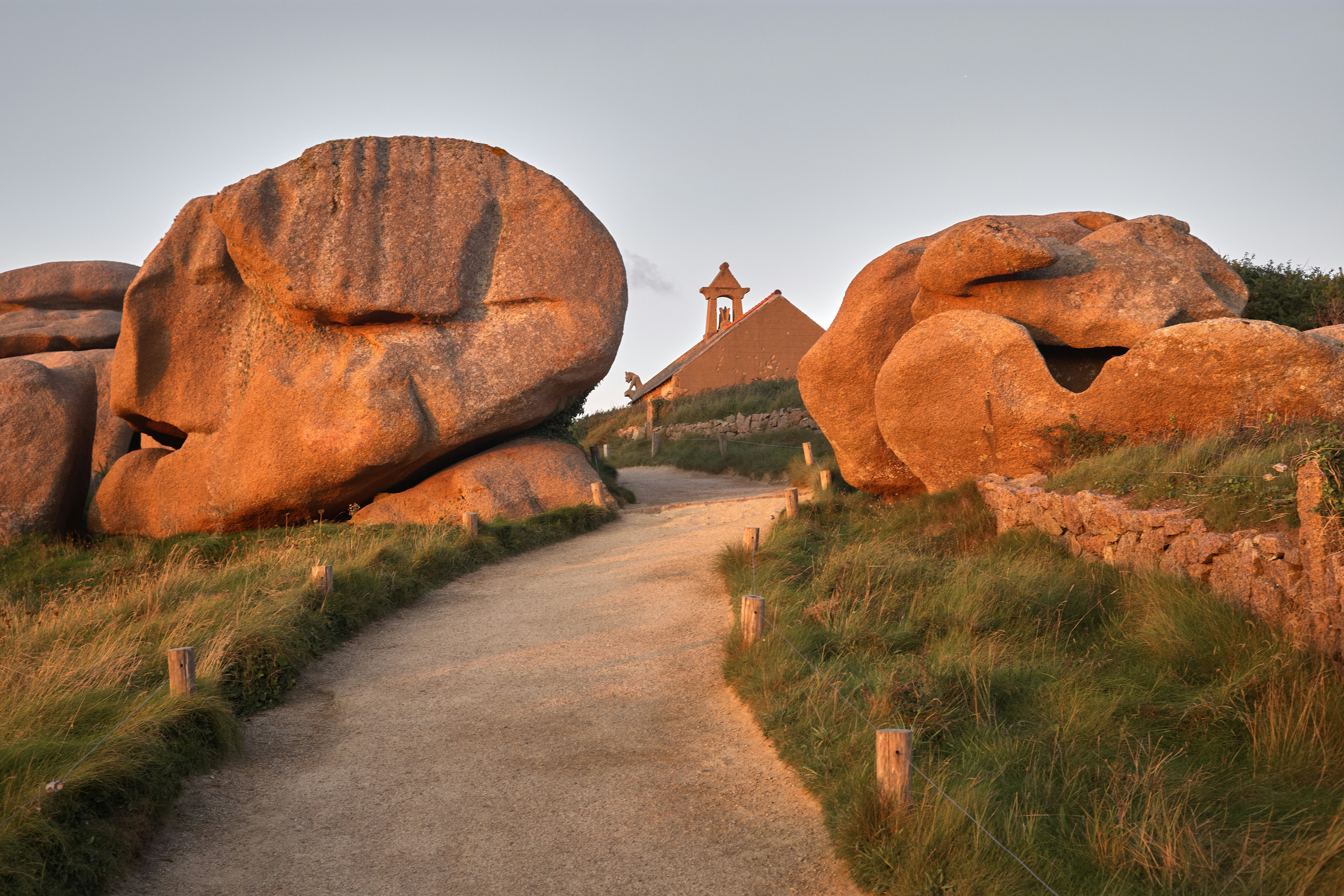 Granite boulders frame a winding path leading to a quaint chapel, bathed in warm evening light.