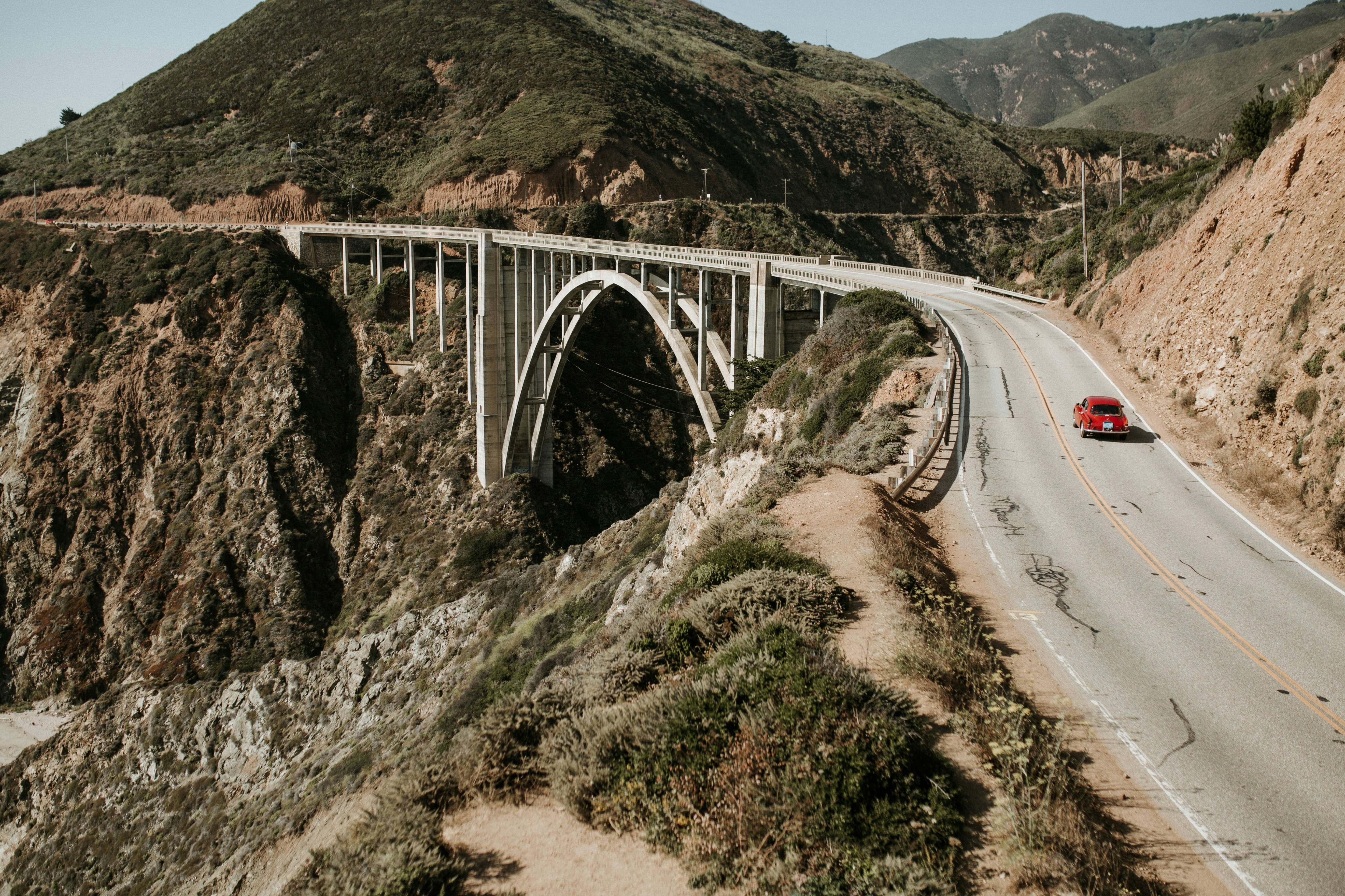 Red car traveling over Bixby Bridge in Big Sur with rugged hills in the background.