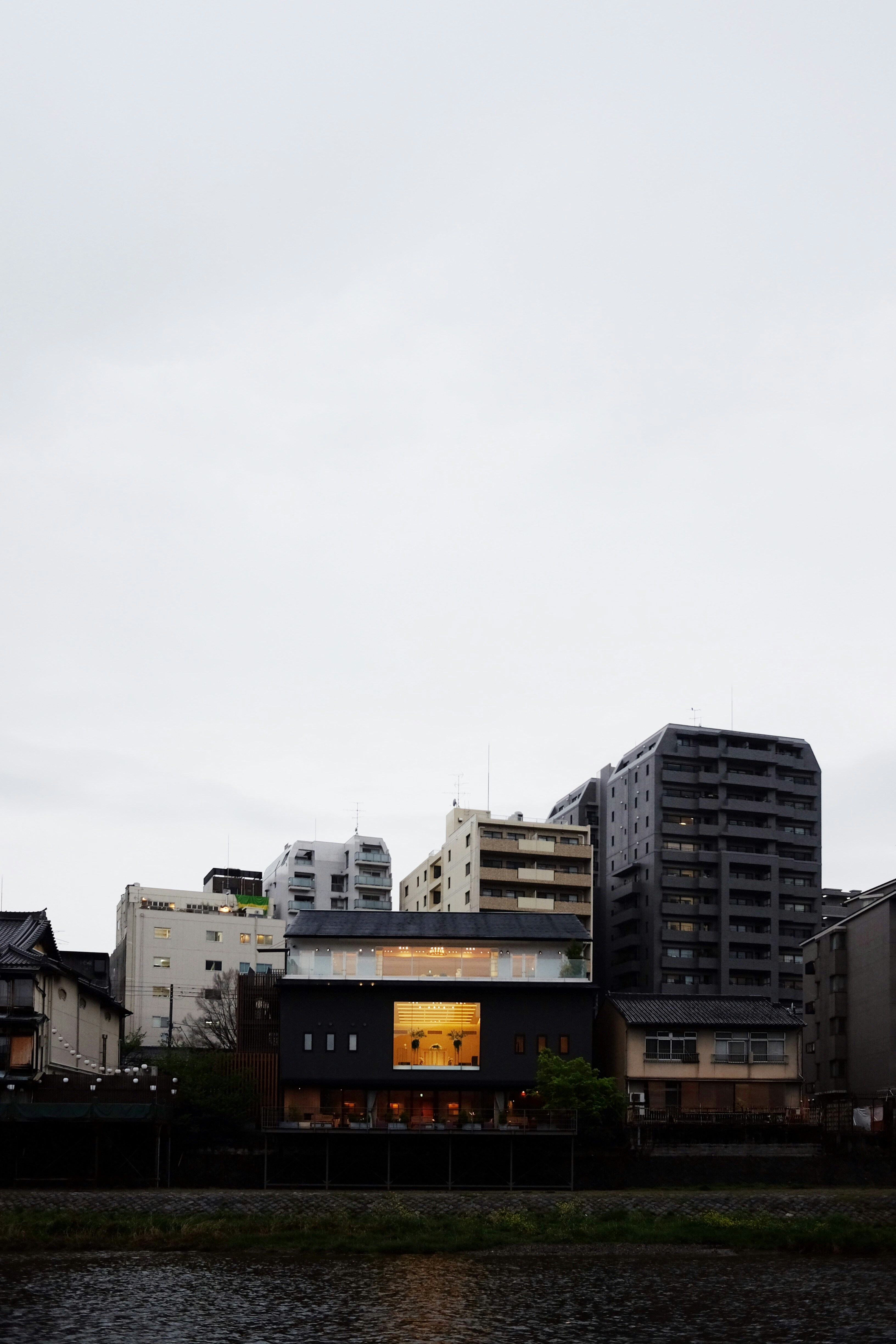 A contemporary building illuminated at dusk contrasts with surrounding high-rises along a riverbank.