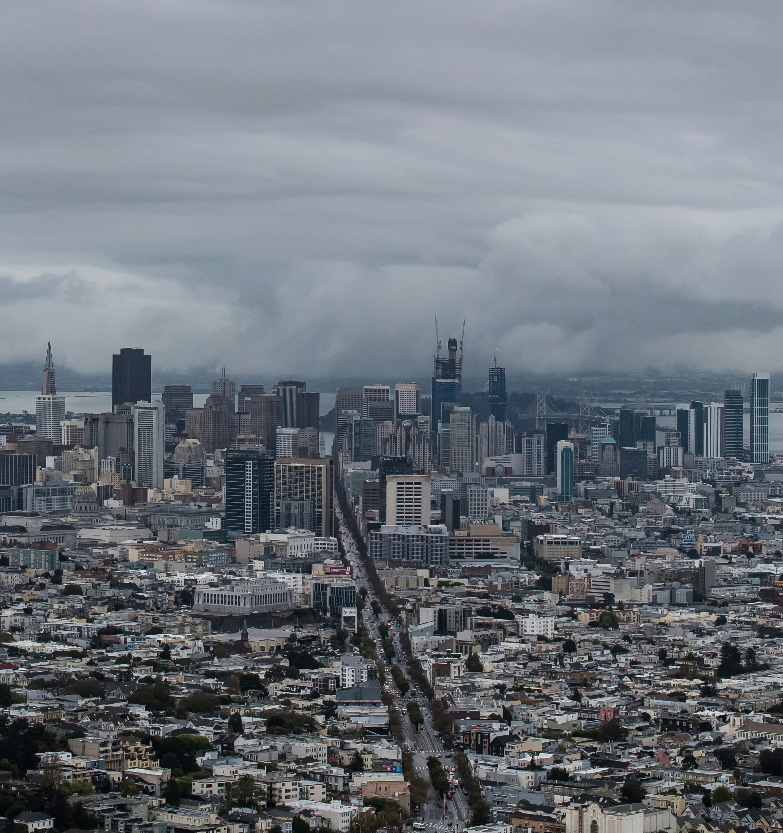 Aerial view of a sprawling cityscape under a blanket of clouds, showcasing the intricate layout of buildings and streets. The skyline features notable structures peeking through the overcast sky.