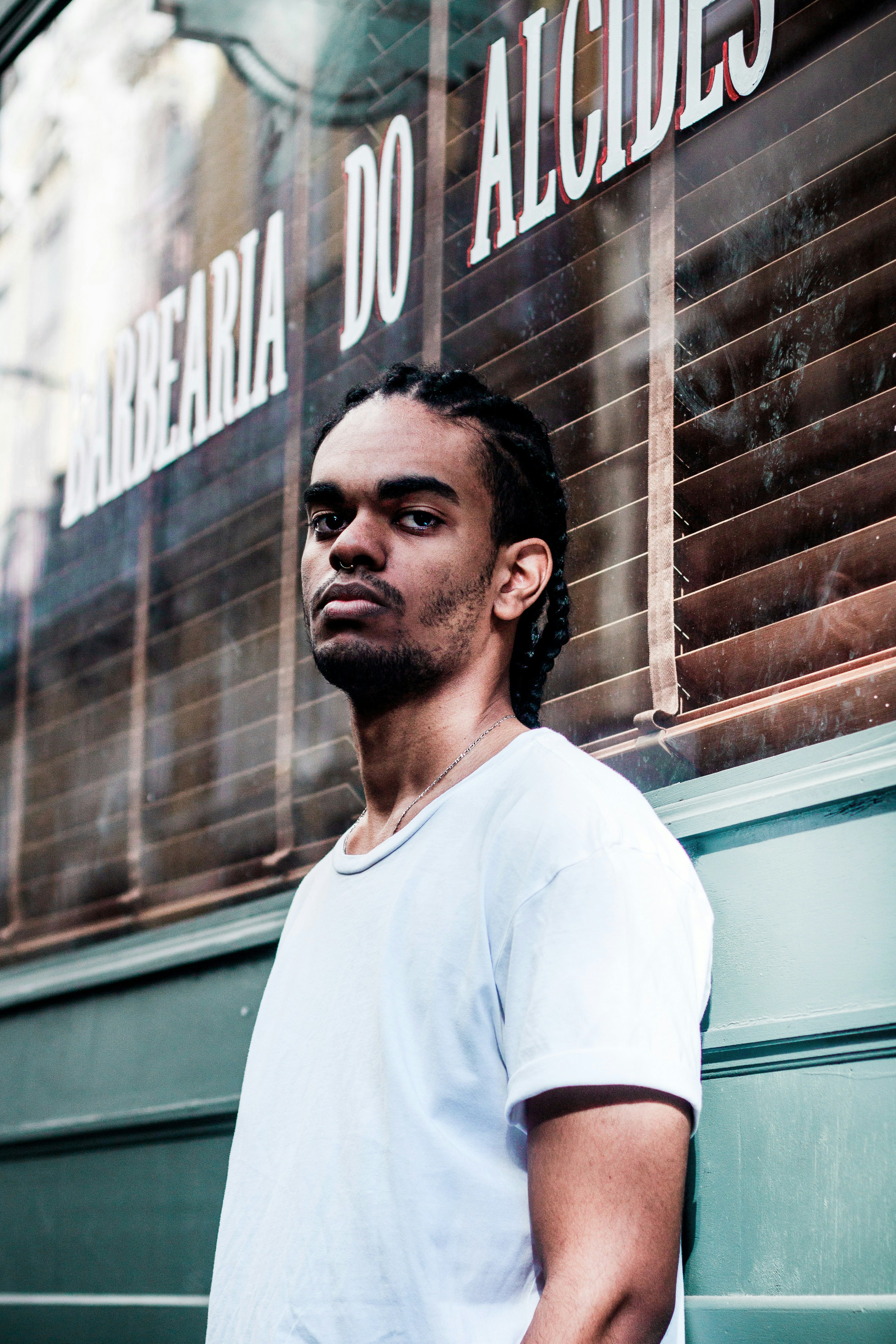 A young man with braided hair stands confidently beside a window with the words 'Barbearia do Alcides' in bold letters.