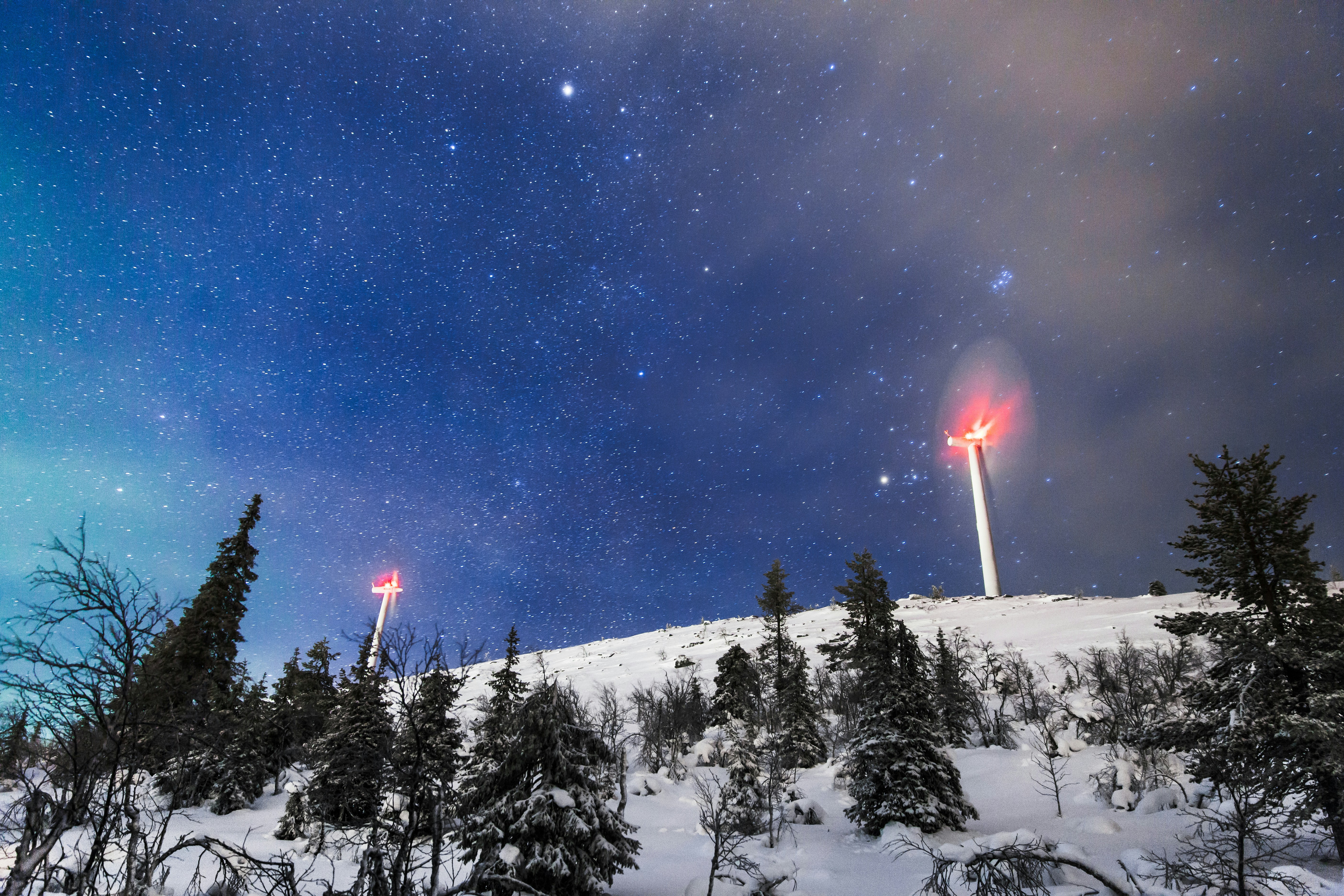 Wind turbines stand tall against a starry night sky, illuminated by distant celestial bodies and soft hues of the aurora. Snow blankets the ground, enhancing the tranquil atmosphere.