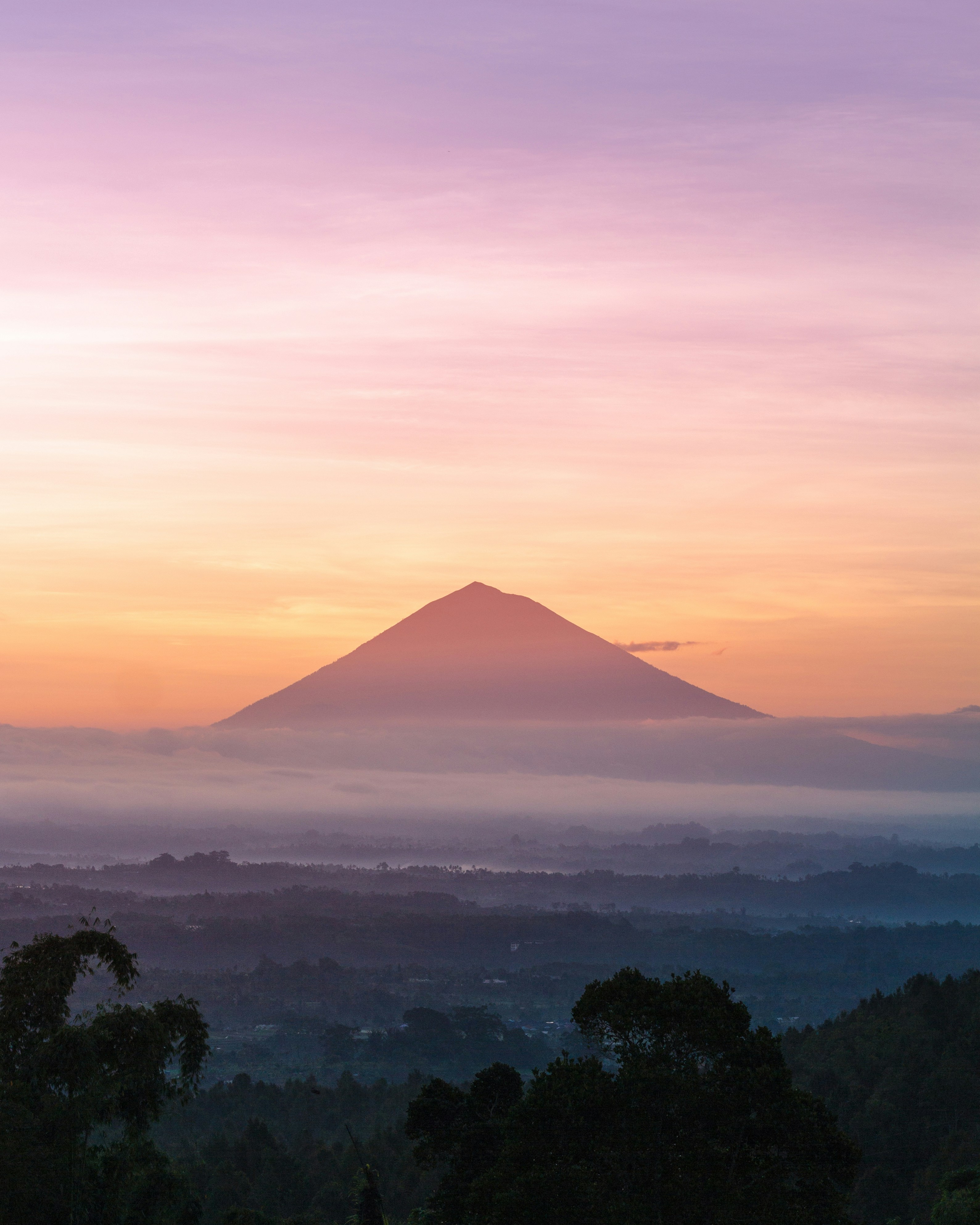 Mountain silhouetted against a vibrant orange and purple sunrise with misty valleys below.