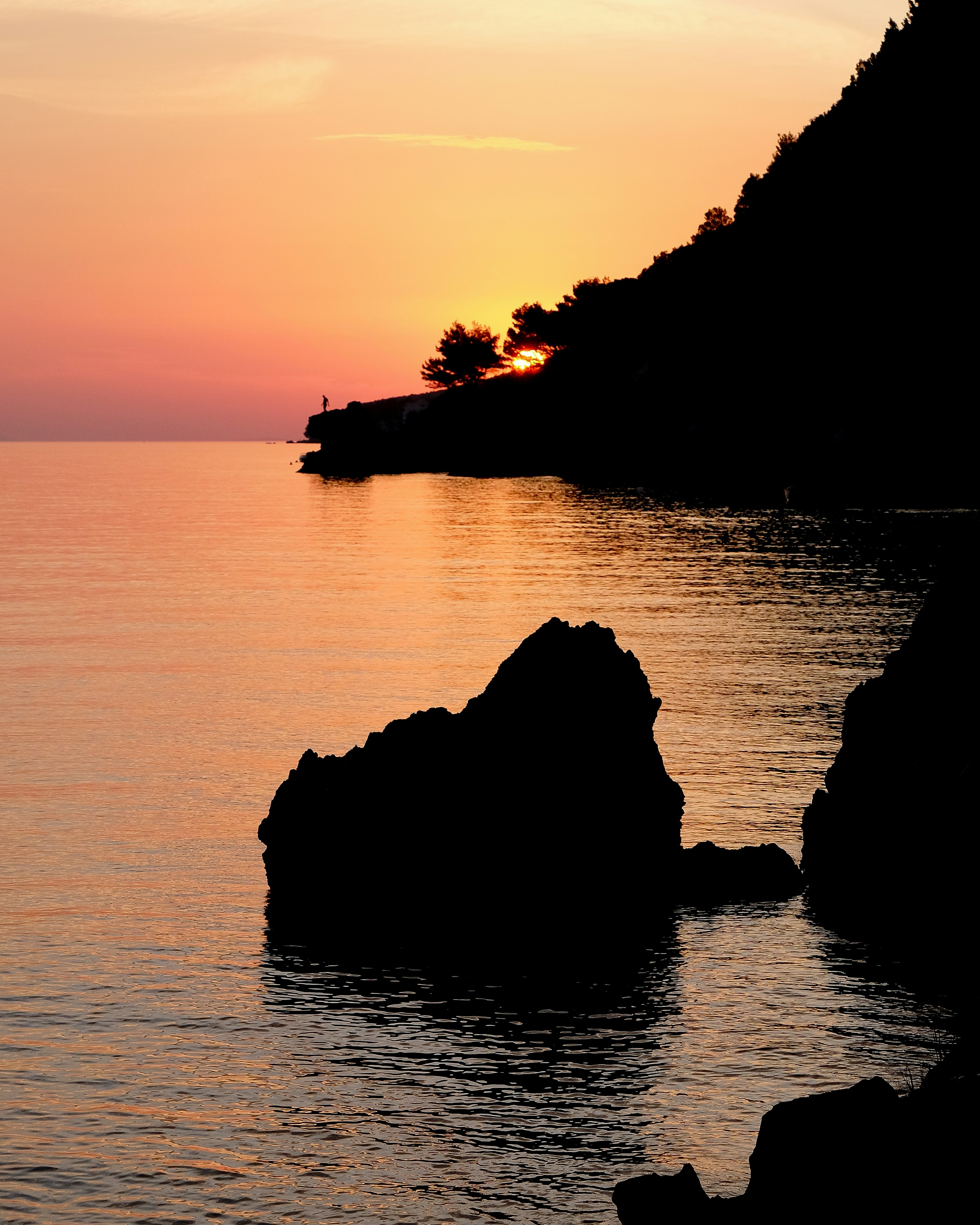Silhouetted rocks and a distant figure against a vibrant sunset over calm waters.