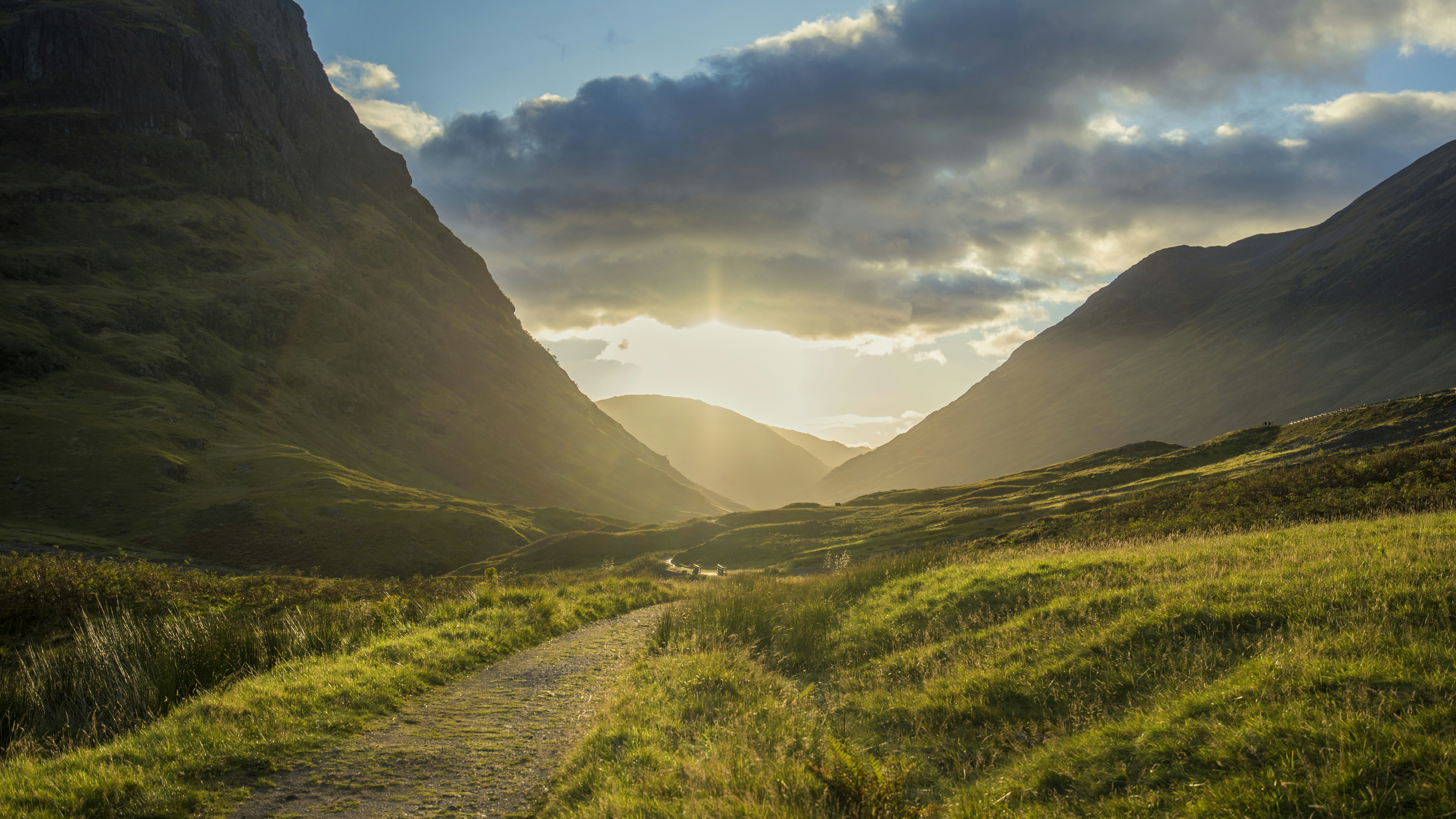 Sunlit valley path winding between shadowed mountains under a dramatic sky.