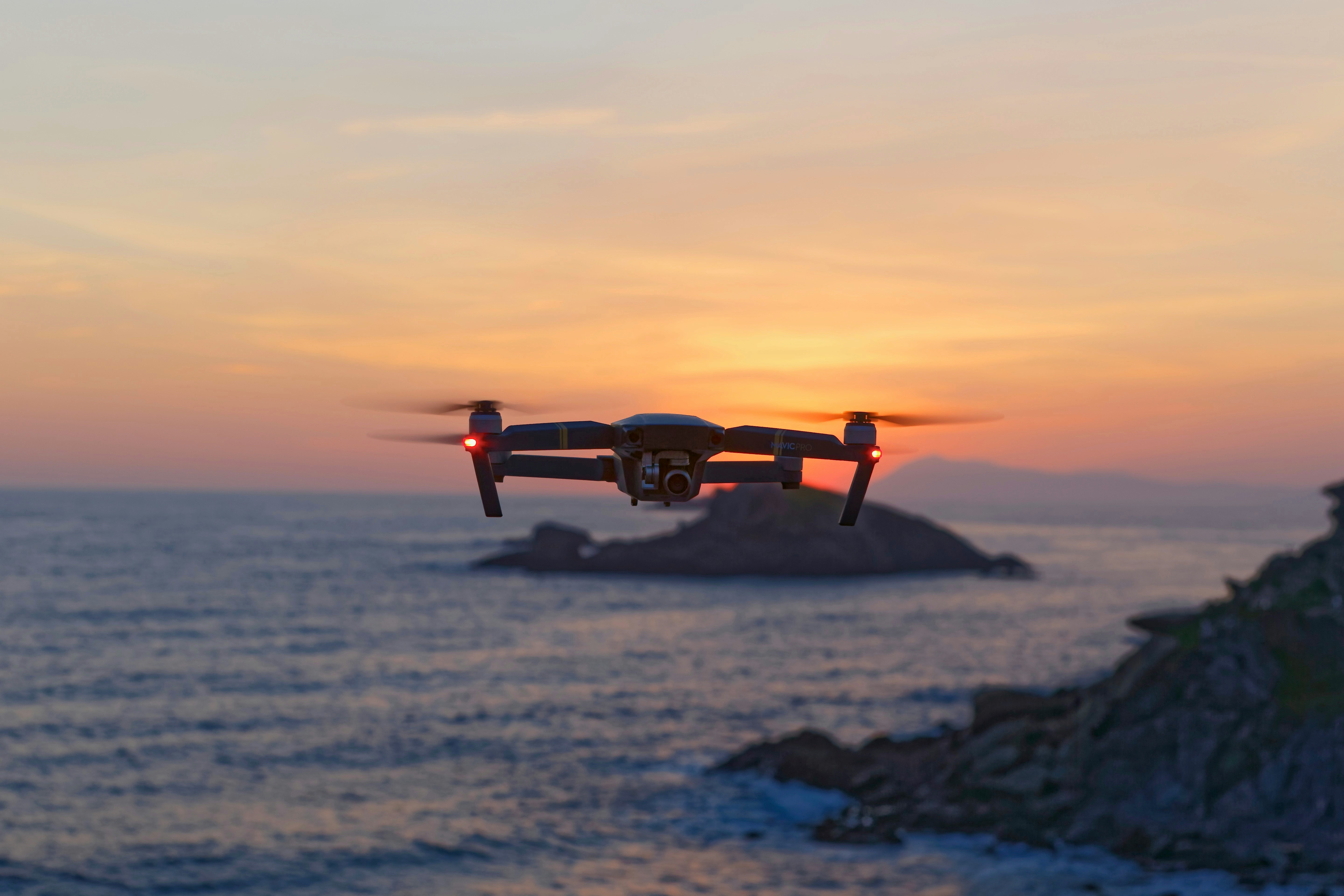Drone hovering gracefully above the ocean at sunset, capturing the serene beauty of the landscape. The warm hues of the sky reflect on the water's surface.