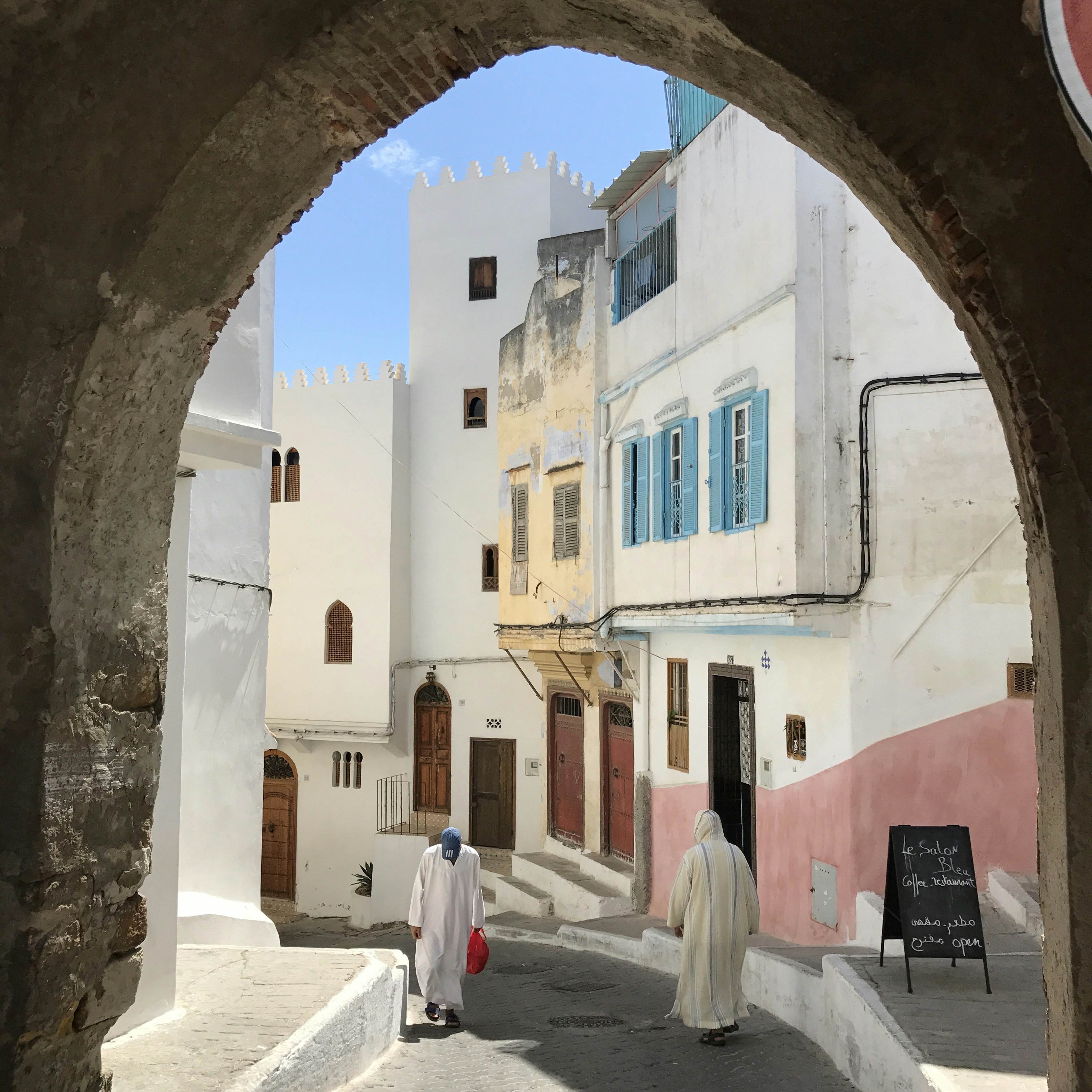Two figures in traditional attire stroll through a historic archway, revealing a vibrant medina with colorful buildings and blue shutters. A sign hints at local offerings.