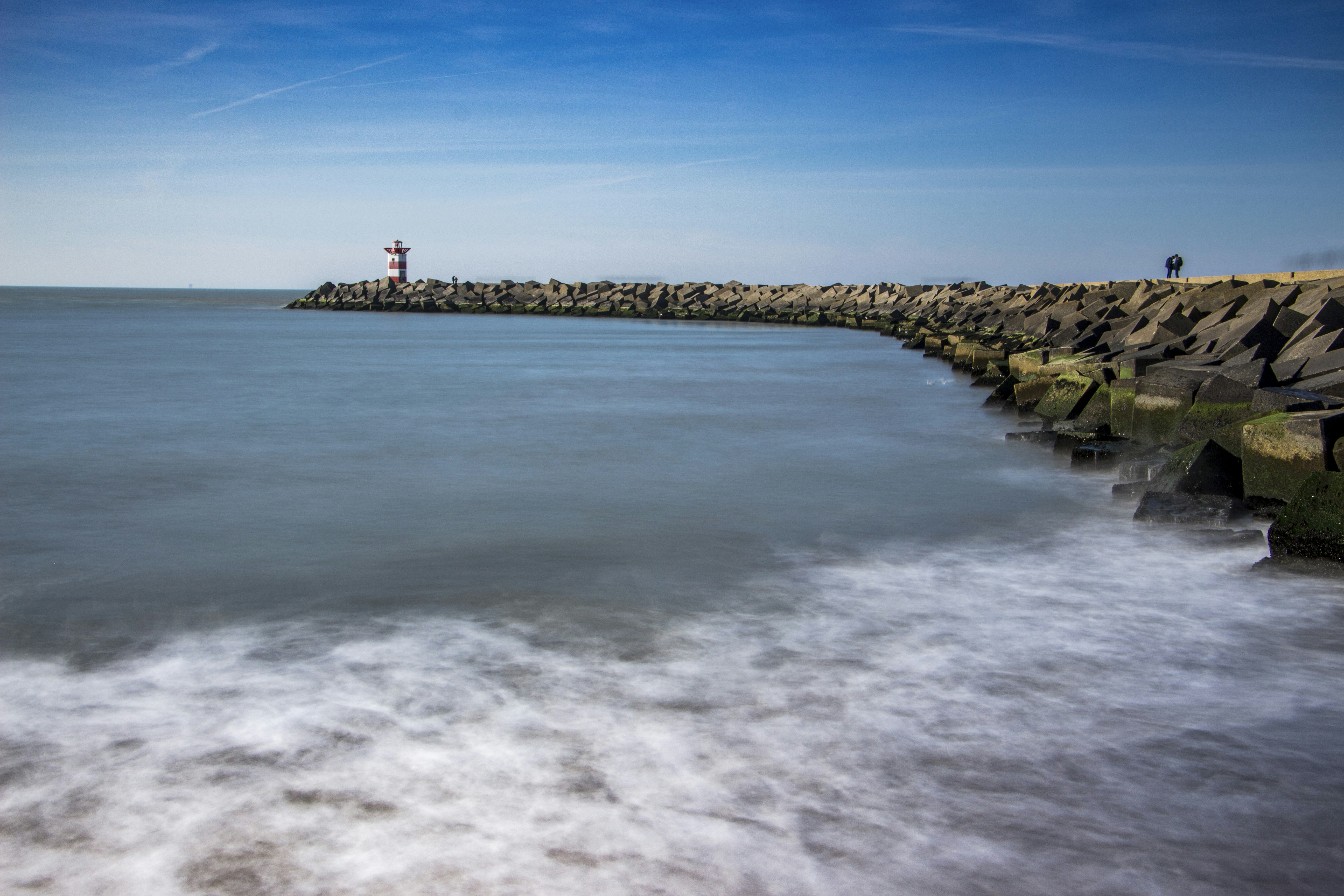Long exposure of waves gently lapping against a stone jetty under a clear blue sky.