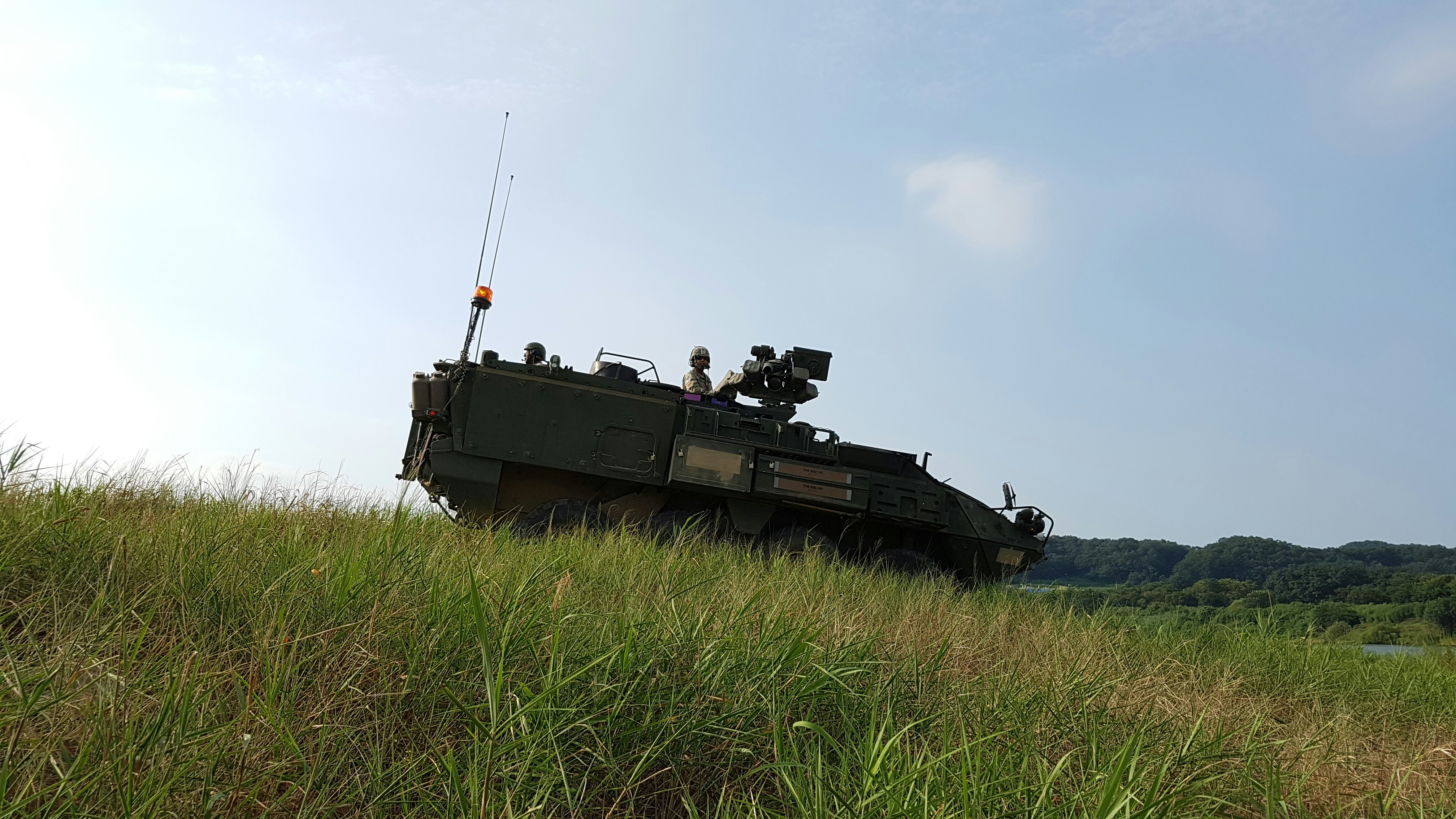Military vehicle traversing tall grass under a clear blue sky.