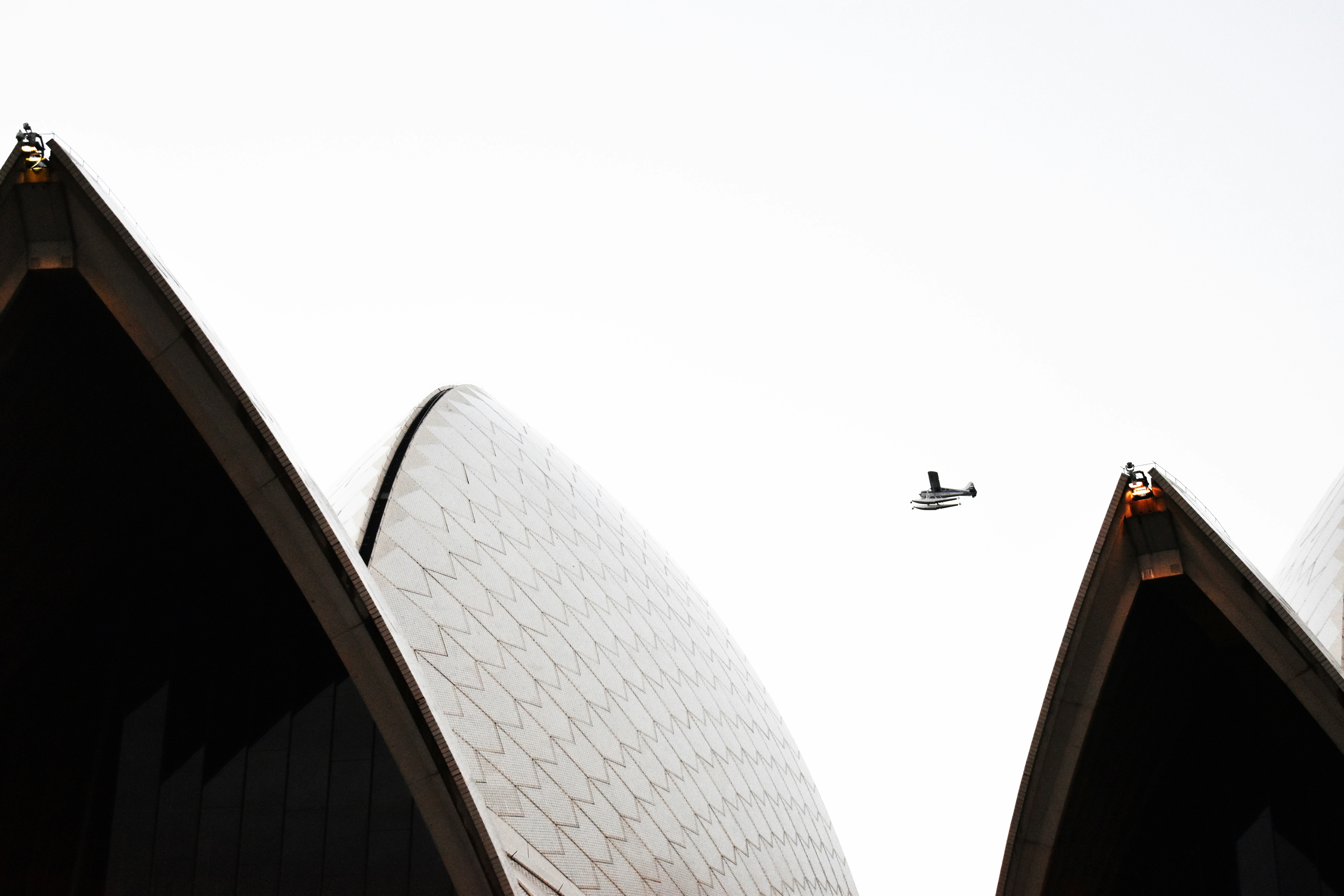 Airplane flying above the curved rooftops of the Sydney Opera House against a clear sky.