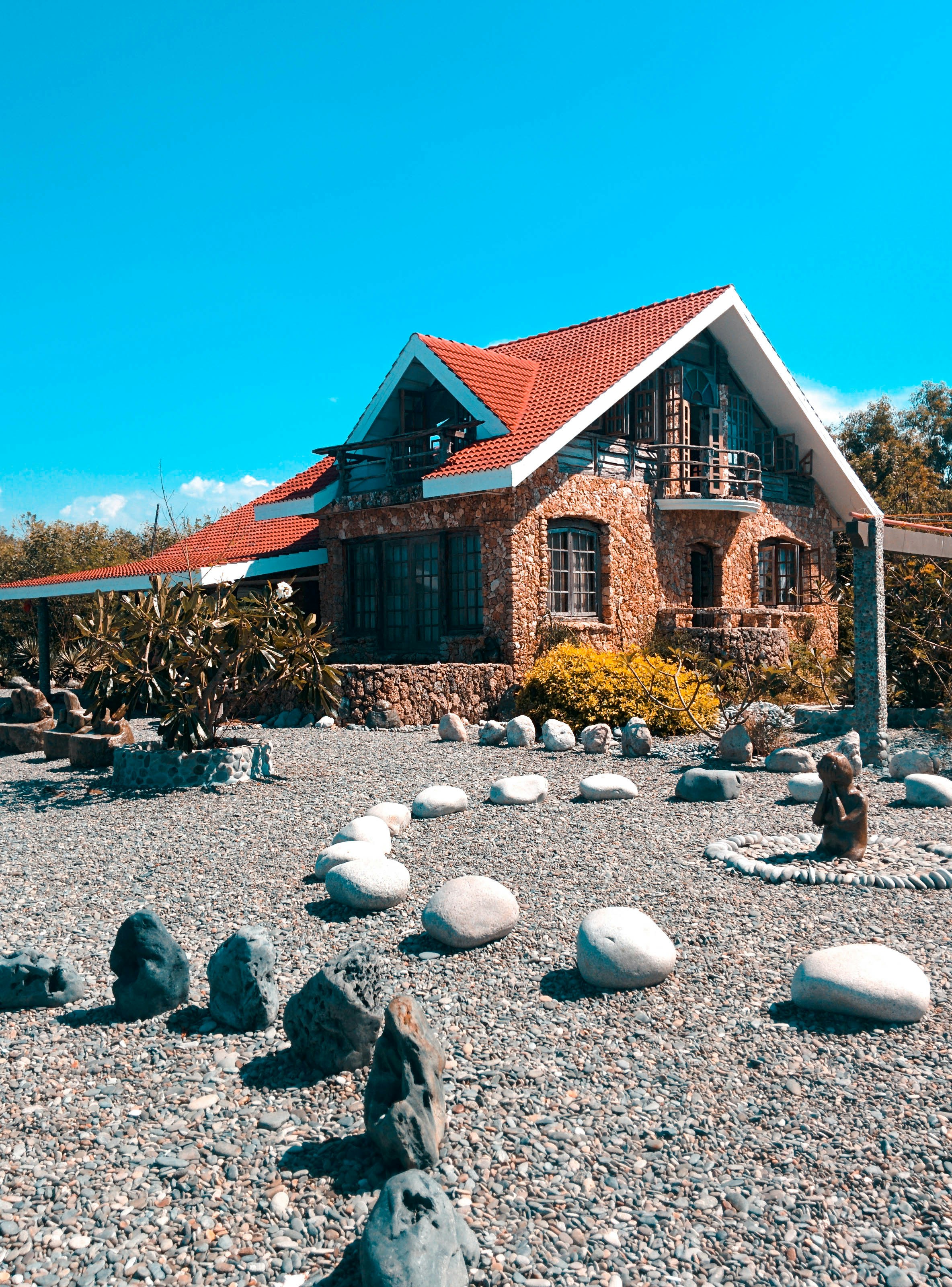 Stone cottage with red roof surrounded by a pebble garden under a bright blue sky.