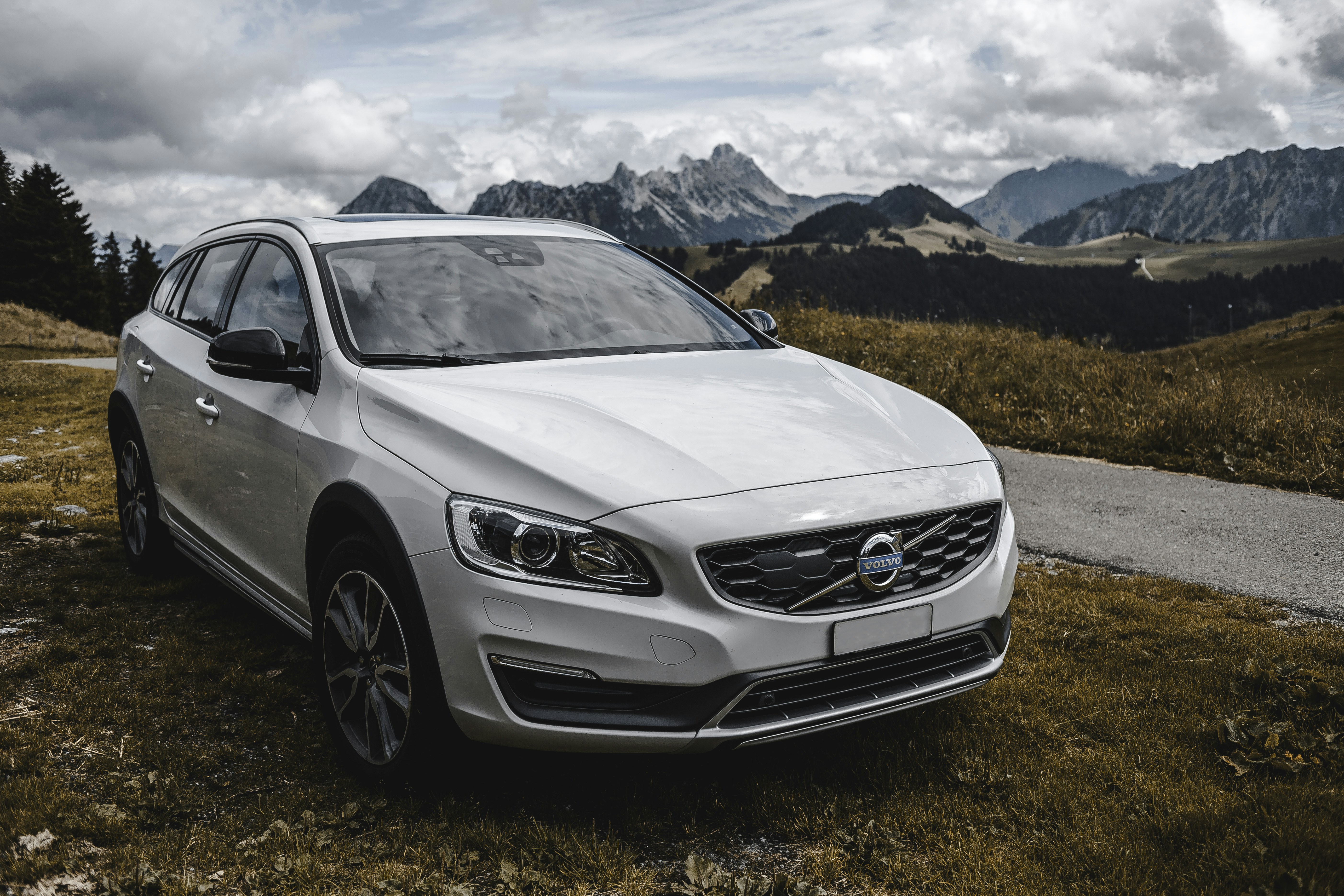 White Volvo station wagon parked on a mountain road with dramatic peaks in the background. The scene conveys a sense of exploration and freedom.