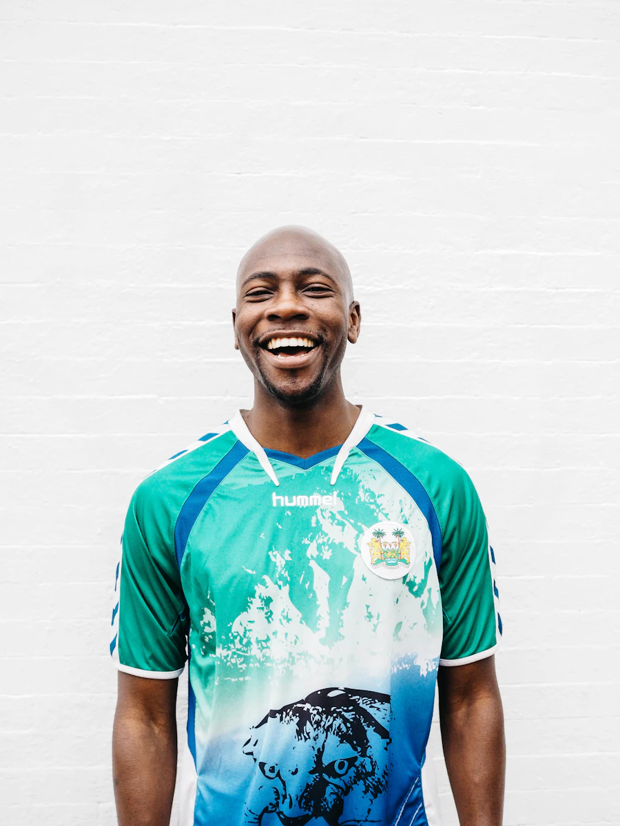 Smiling man standing against a white textured wall, wearing a teal-and-blue sports jersey. His face is centered and brightly lit.