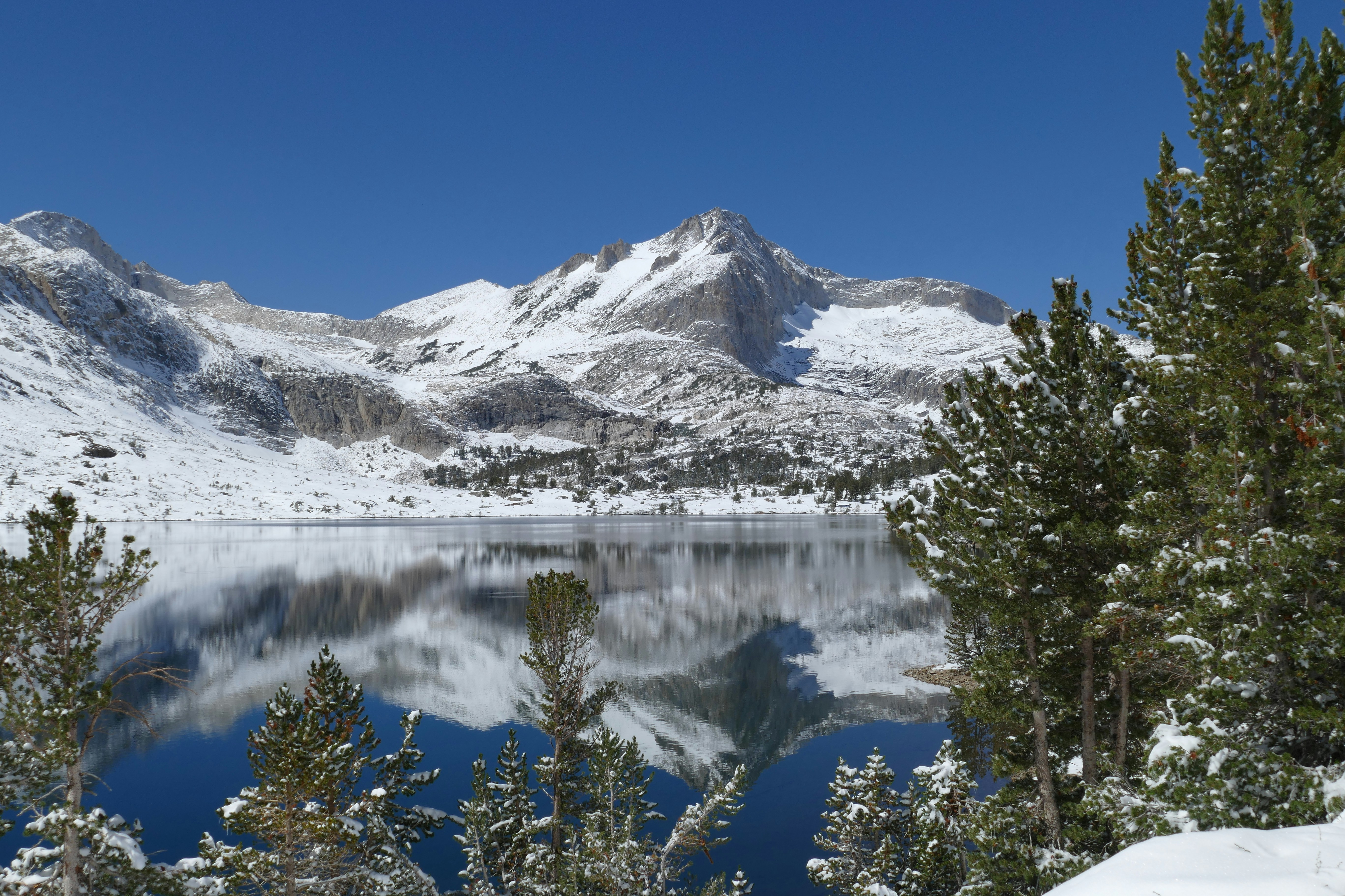 Snowy mountains rise above a calm alpine lake, their reflection creating a near-perfect mirror under a clear blue sky. Evergreen pines frame the foreground.