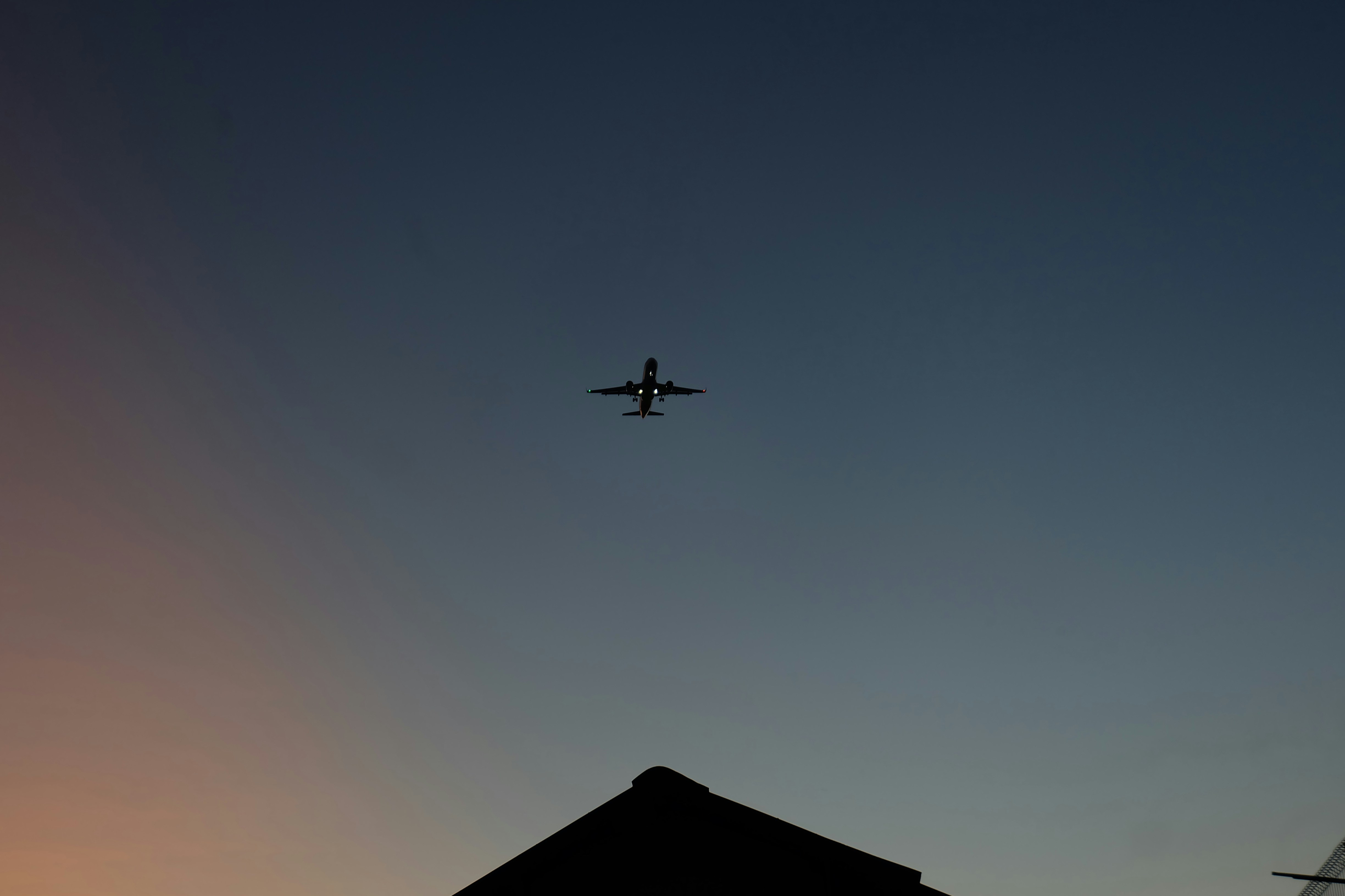 Airplane descending against a twilight sky, silhouetted by a rooftop. The gradient of colors hints at the day's end.
