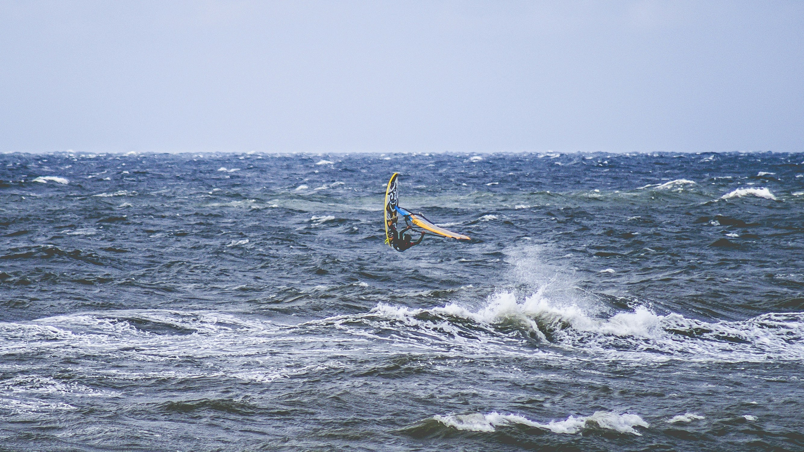 Windsurfer soars above choppy ocean as spray trails, pushed by a gusty wind over breaking waves.