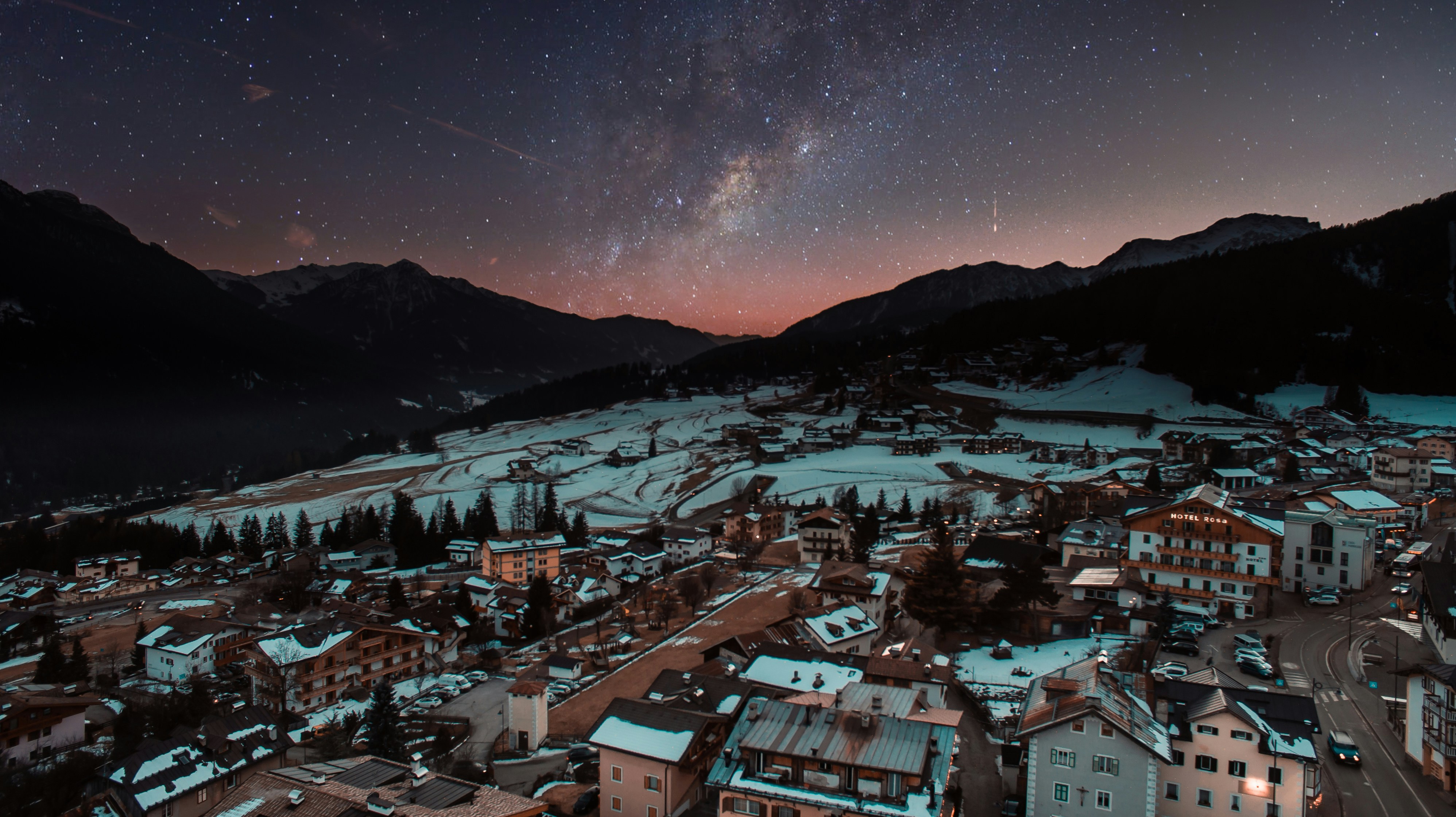 Snow-covered village nestled in a valley under a starry night sky with distant mountains silhouetted against the horizon.