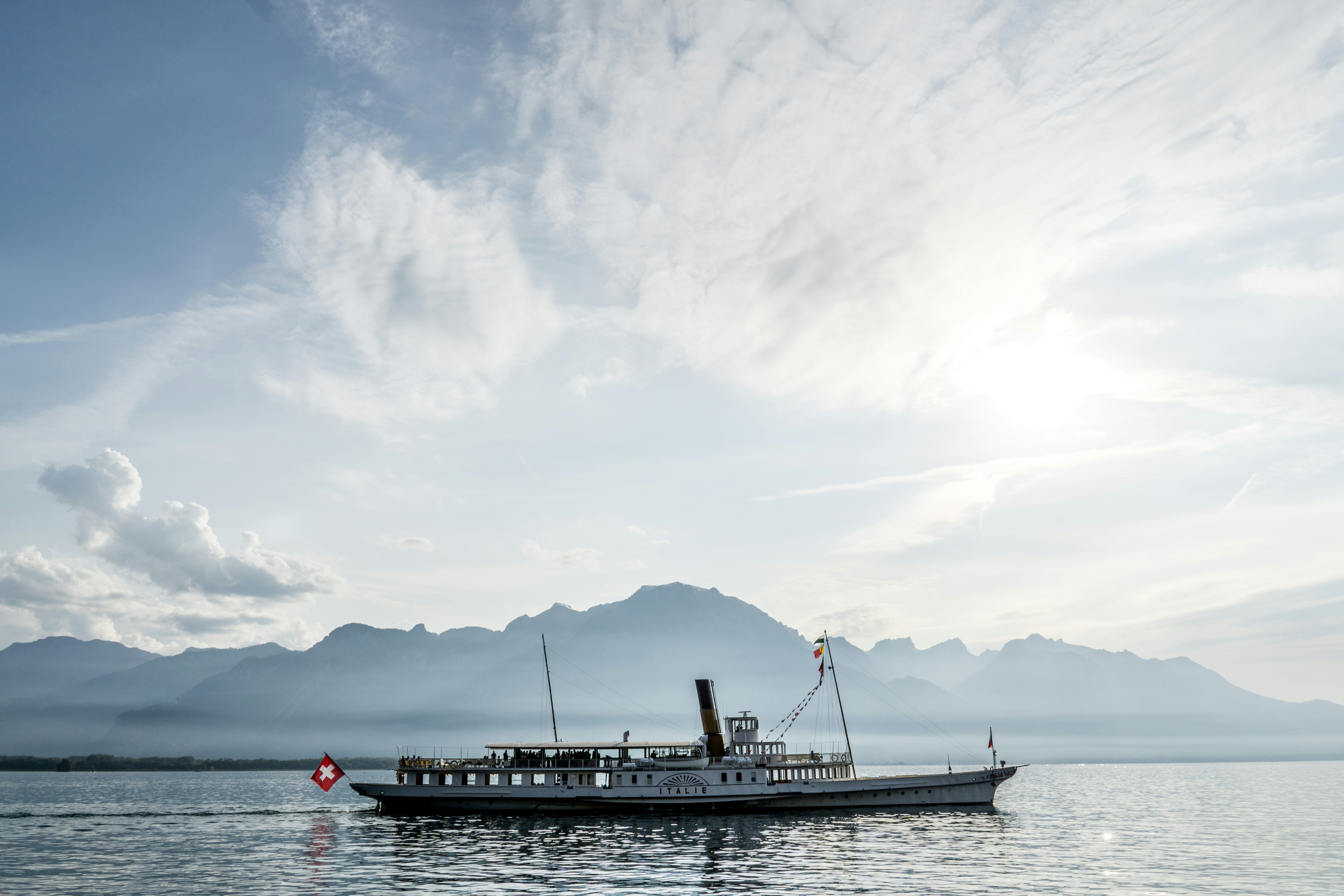 Vintage paddle steamer gliding across calm waters with a mountainous backdrop under a cloudy sky.
