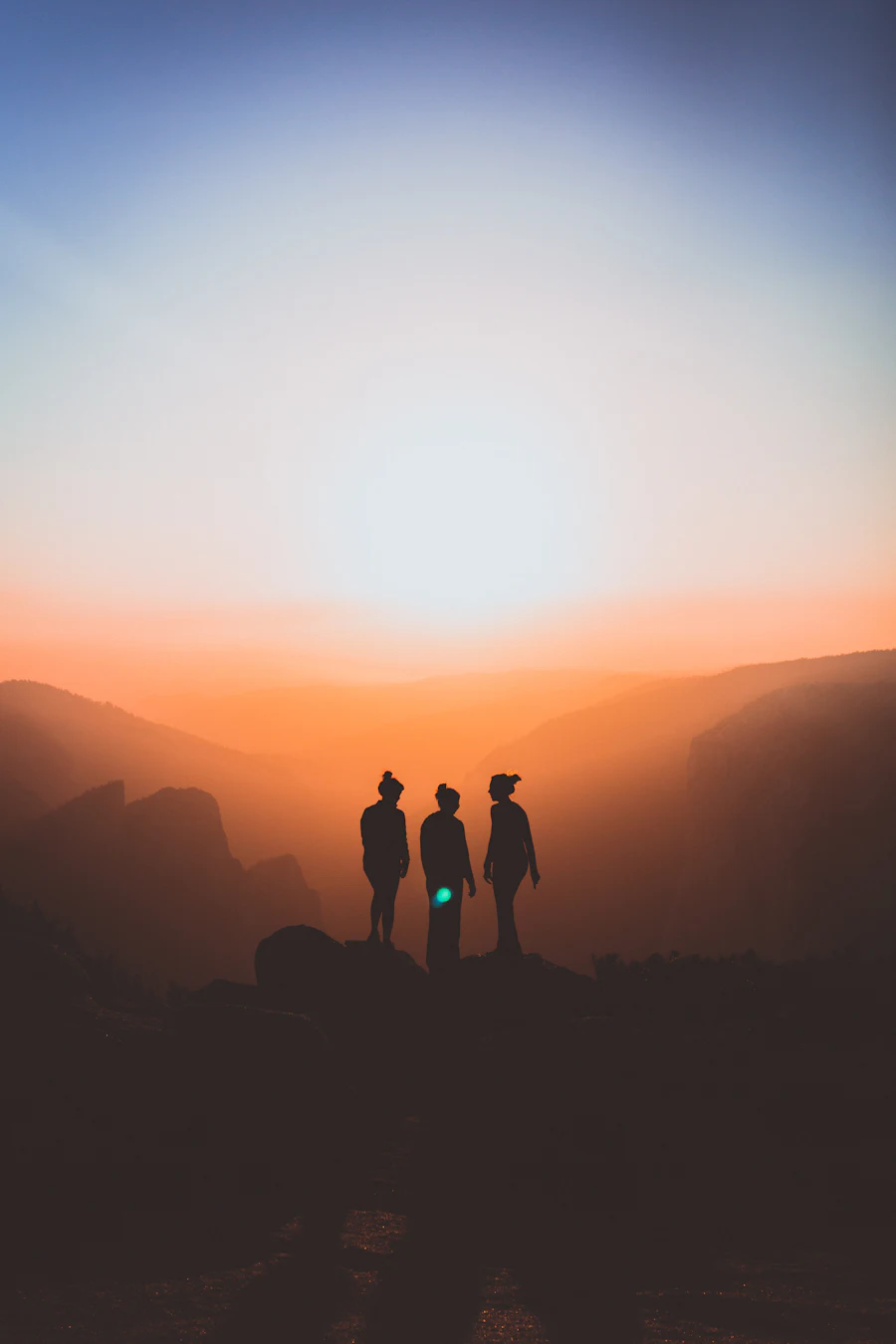 Three silhouetted hikers stand on rocky terrain above layered mountain ridges, with a warm orange sunrise fading into a blue sky. A small lens flare glows near the center.