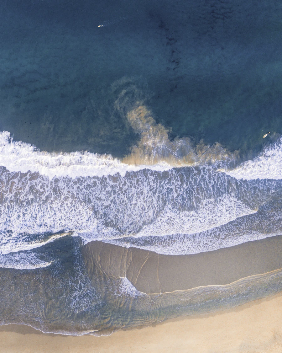 A drone captures turquoise water meeting foamy waves along a sandy beach, creating a vivid border between sea and shore. The image reveals a coastal shoreline from above, with natural color gradations and gentle wave patterns.