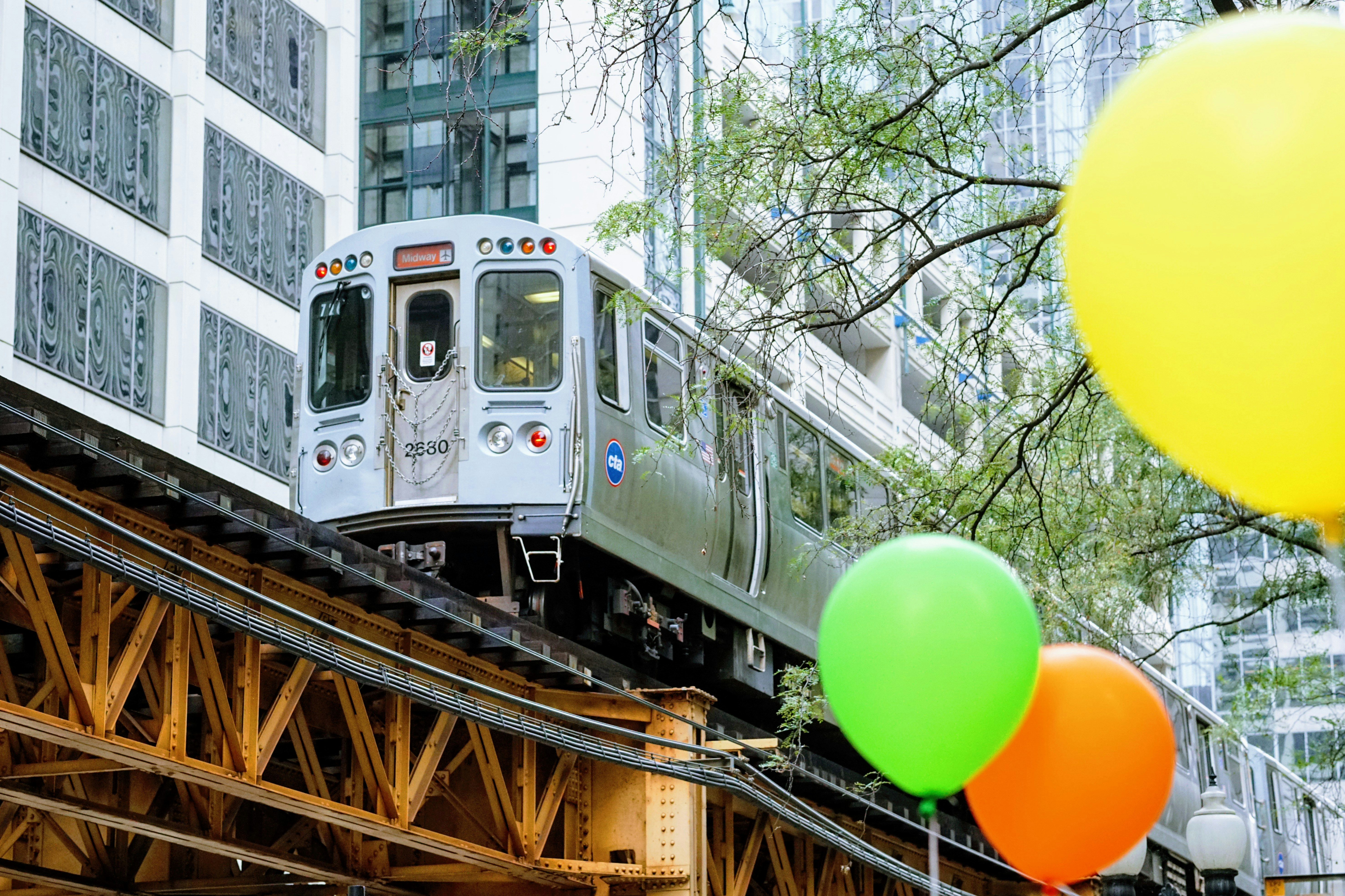 A silver train glides along an elevated track, framed by colorful balloons in the foreground and modern buildings in the background.