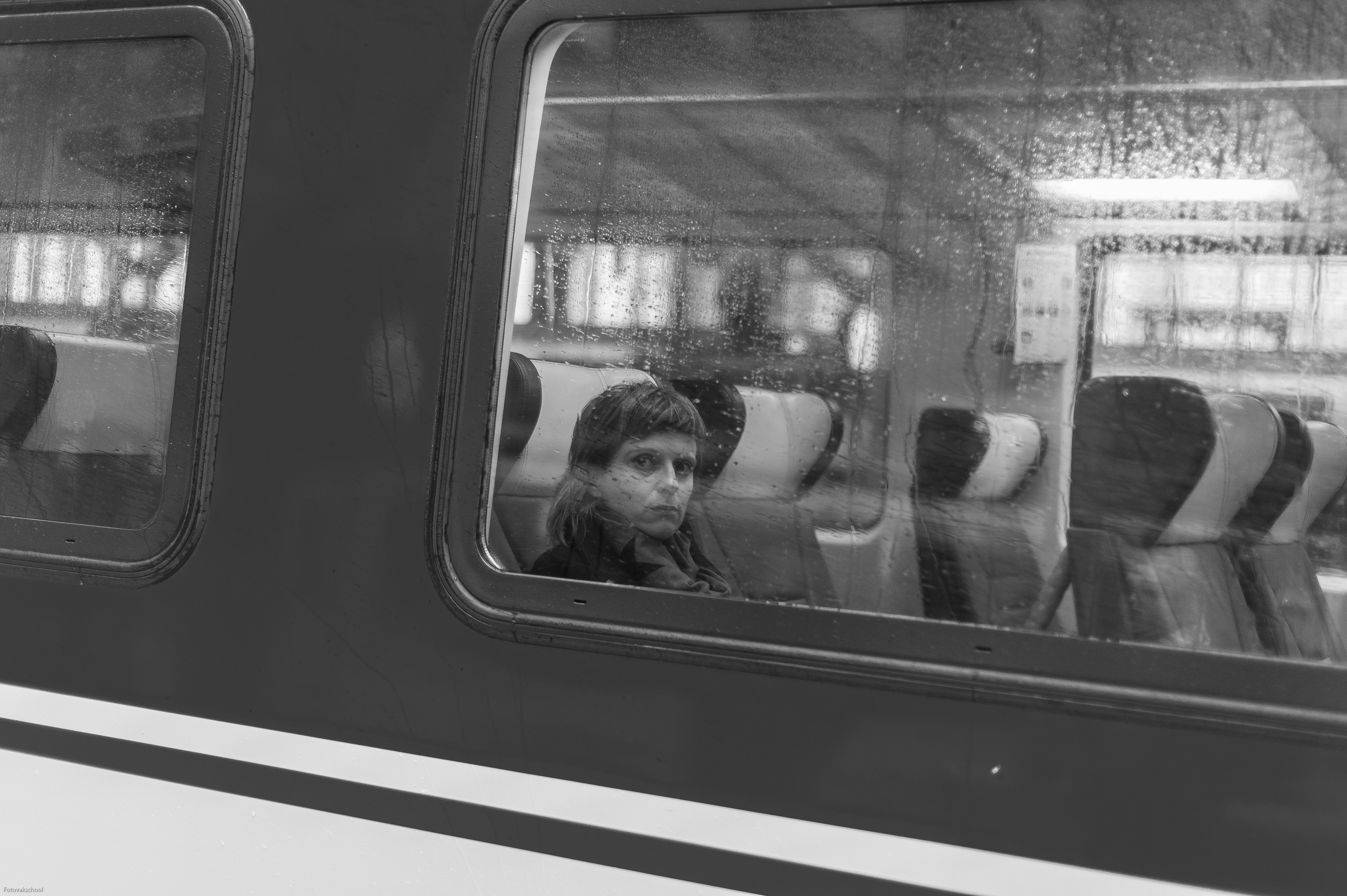 A contemplative figure gazes through a rain-speckled train window, surrounded by empty seats. The scene captures a moment of introspection amidst urban transit.