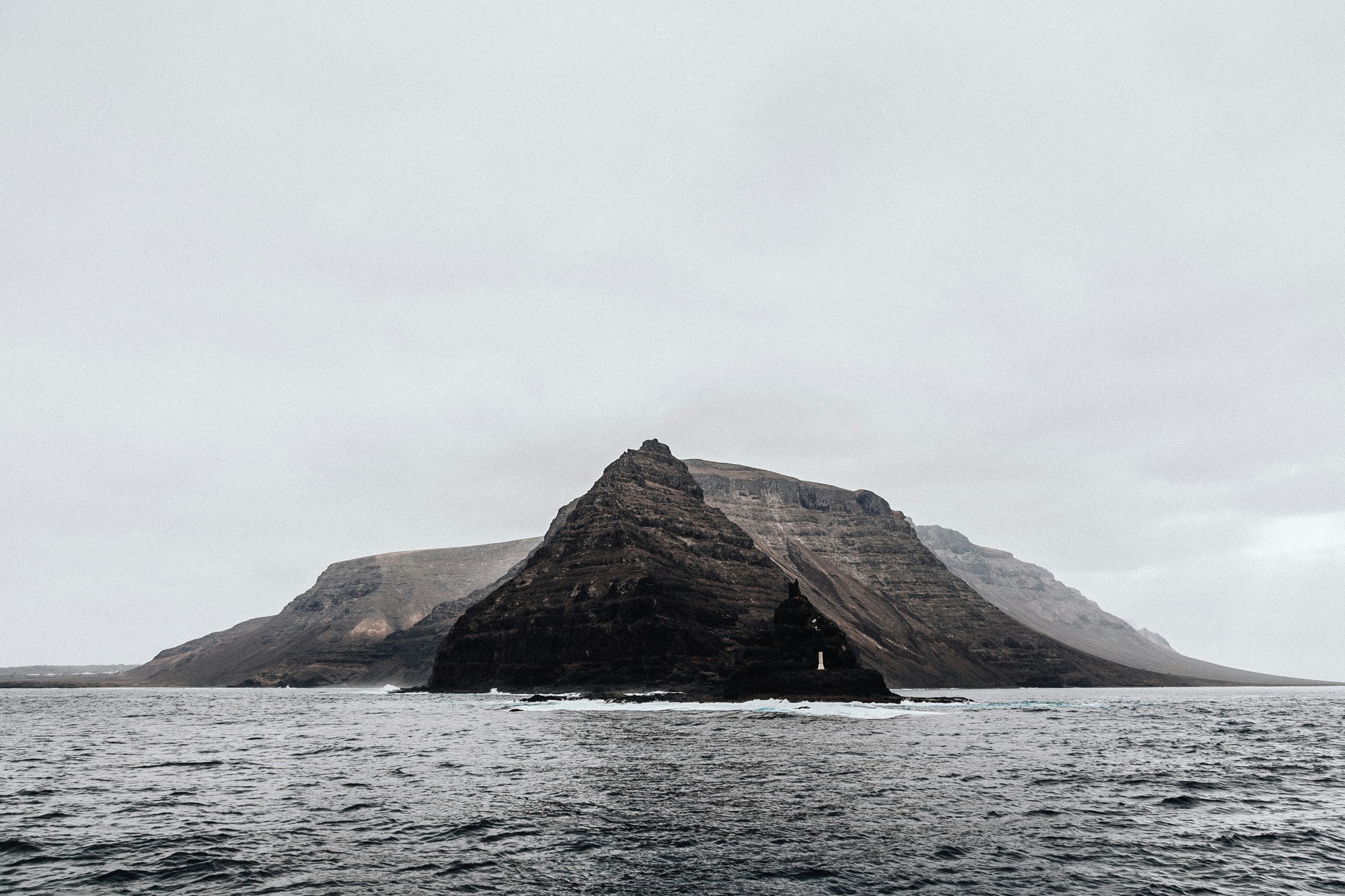 A rugged island silhouette rises from the ocean under a cloudy sky, hinting at the mysteries of the deep. The lighthouse stands as a solitary beacon against the dramatic landscape.