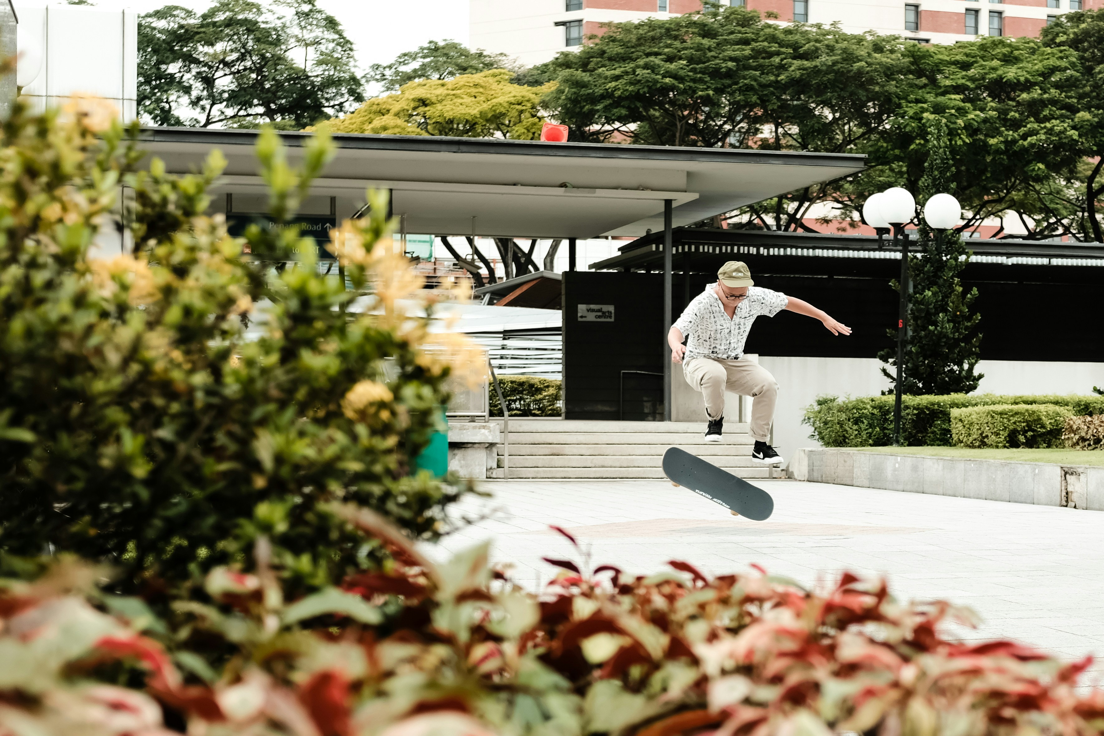 Skateboarder performing a flip trick in a city plaza surrounded by greenery.