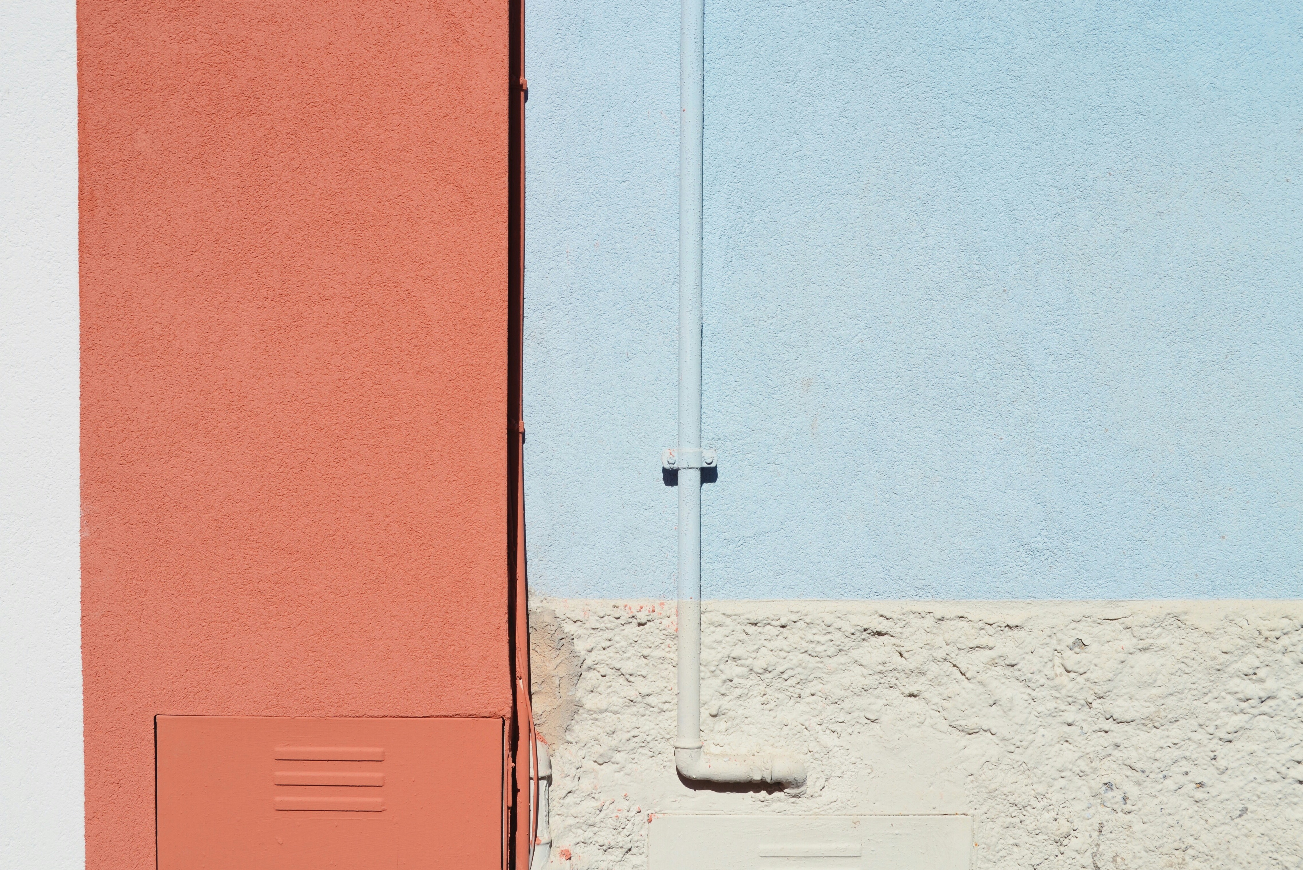 A minimalist composition featuring a vibrant red wall juxtaposed with a soft blue surface, accented by a white pipe and textured lower section.