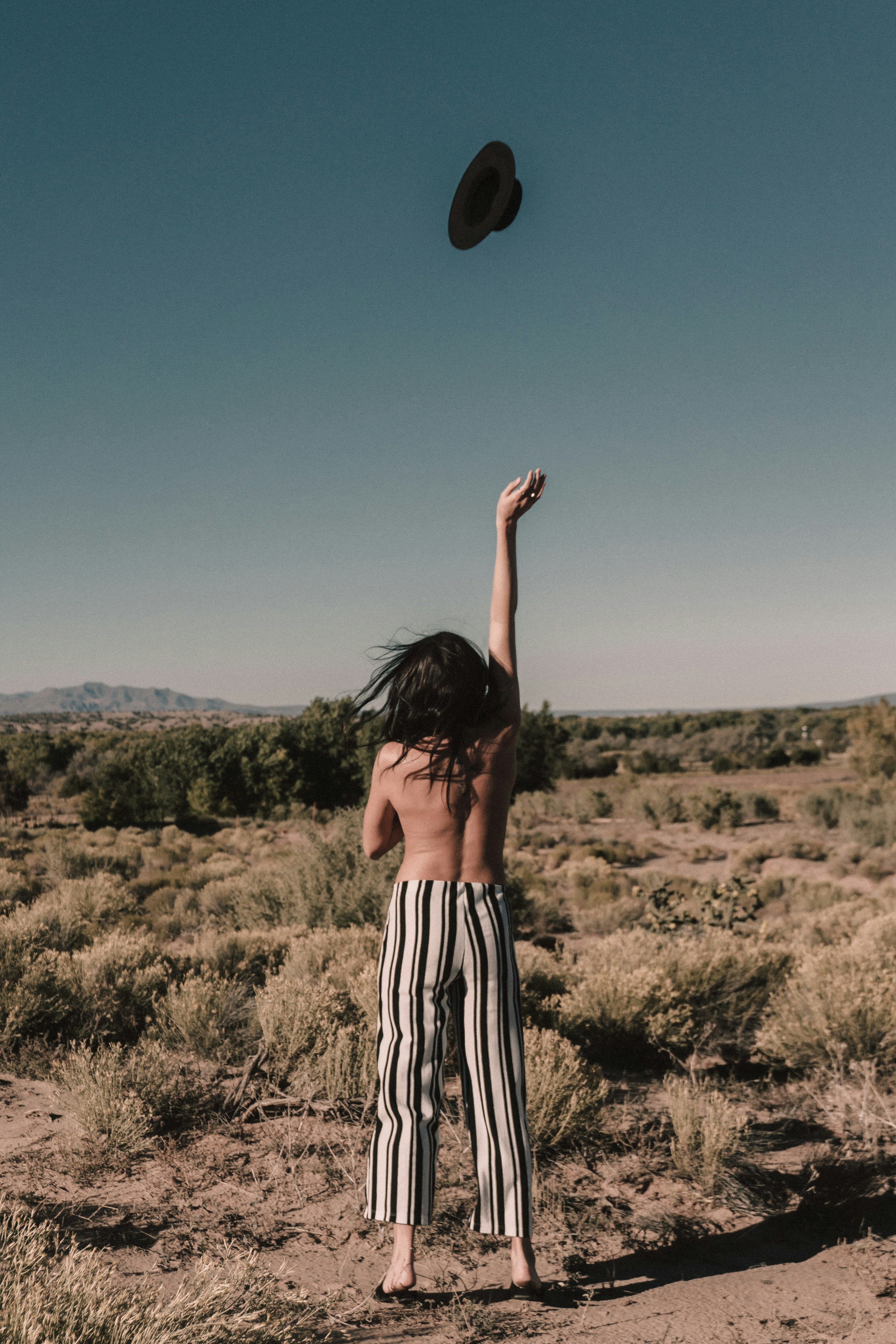 Person with long hair in striped pants tossing a black hat into the air over a desert landscape.