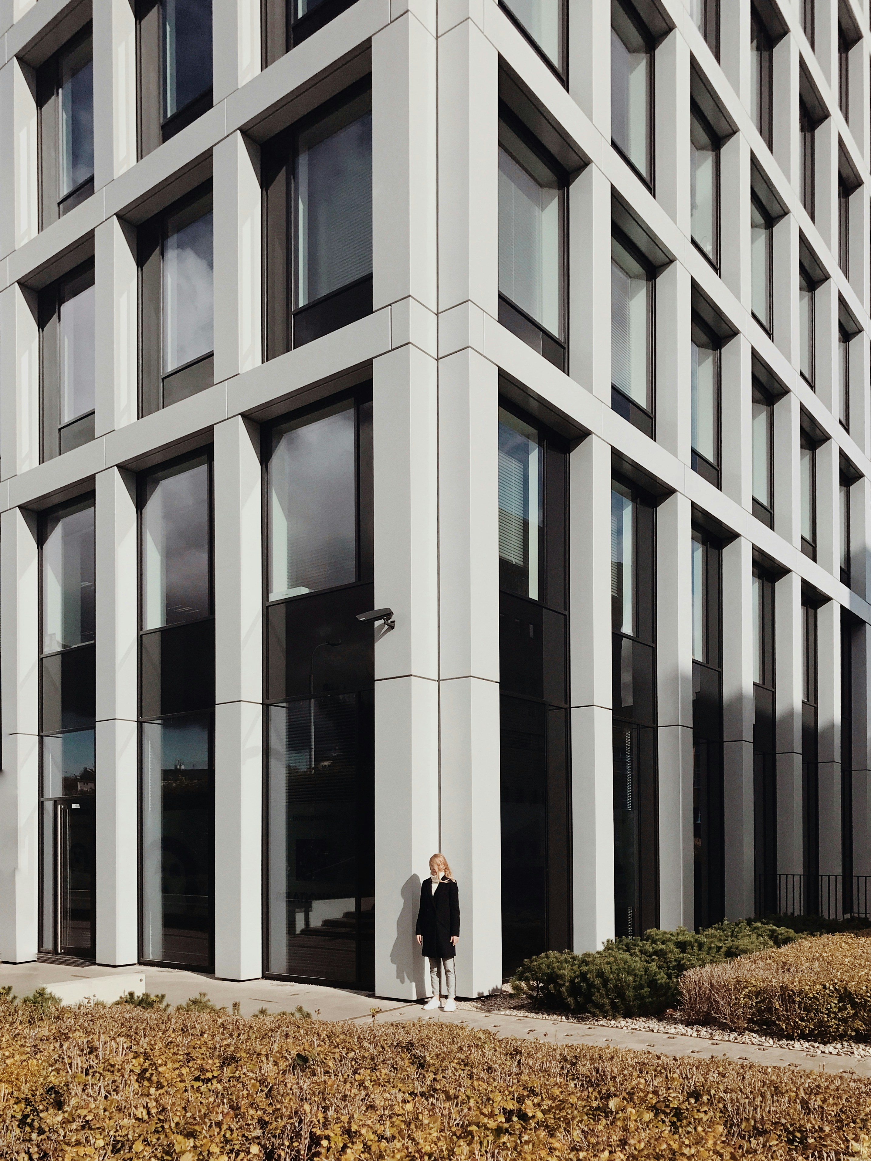 A person stands beside a modern architectural structure, showcasing sharp lines and reflective surfaces. The surrounding greenery adds a touch of nature to the urban scene.