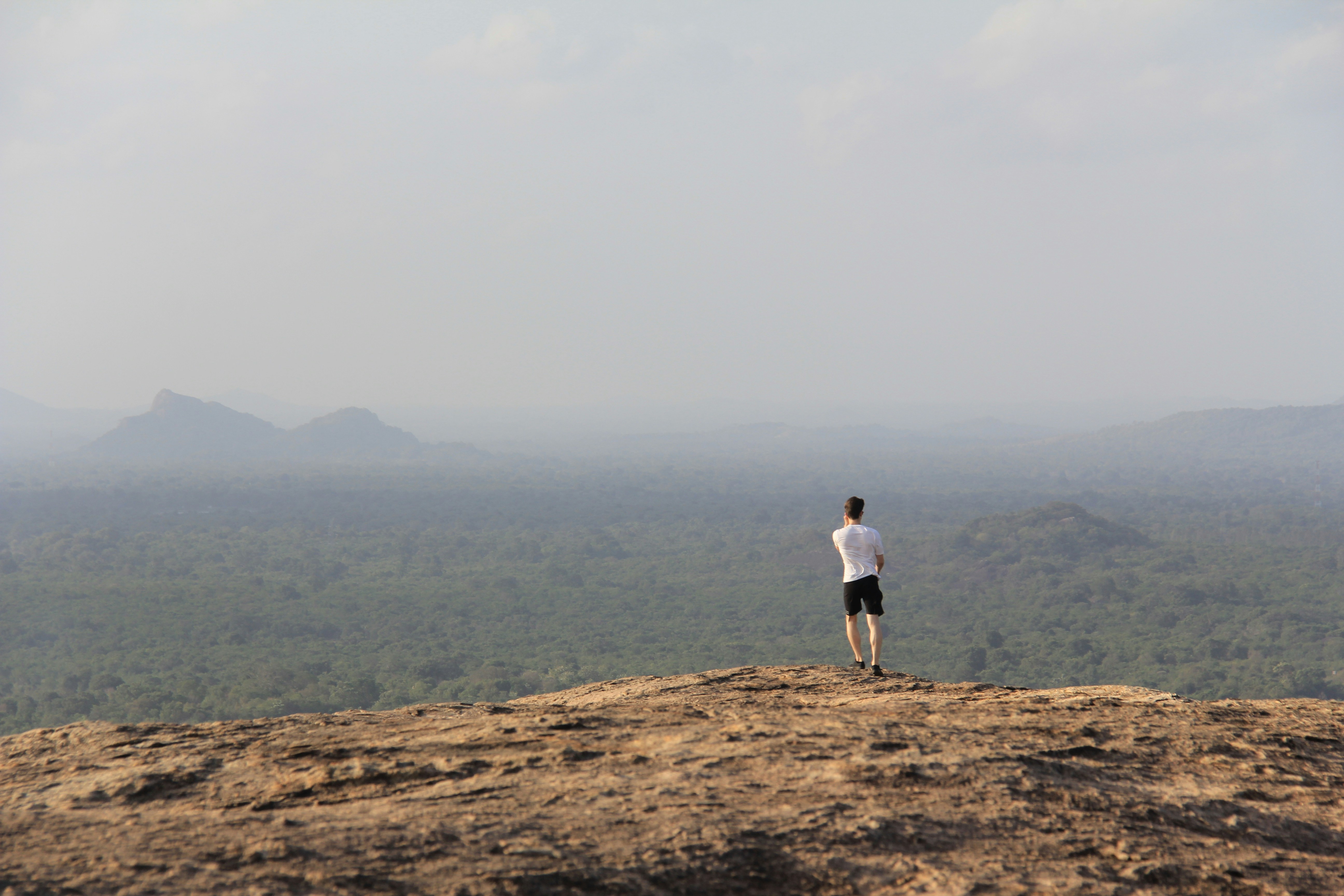 Person in a white T-shirt stands on a rocky mountain peak overlooking a vast, hazy landscape.