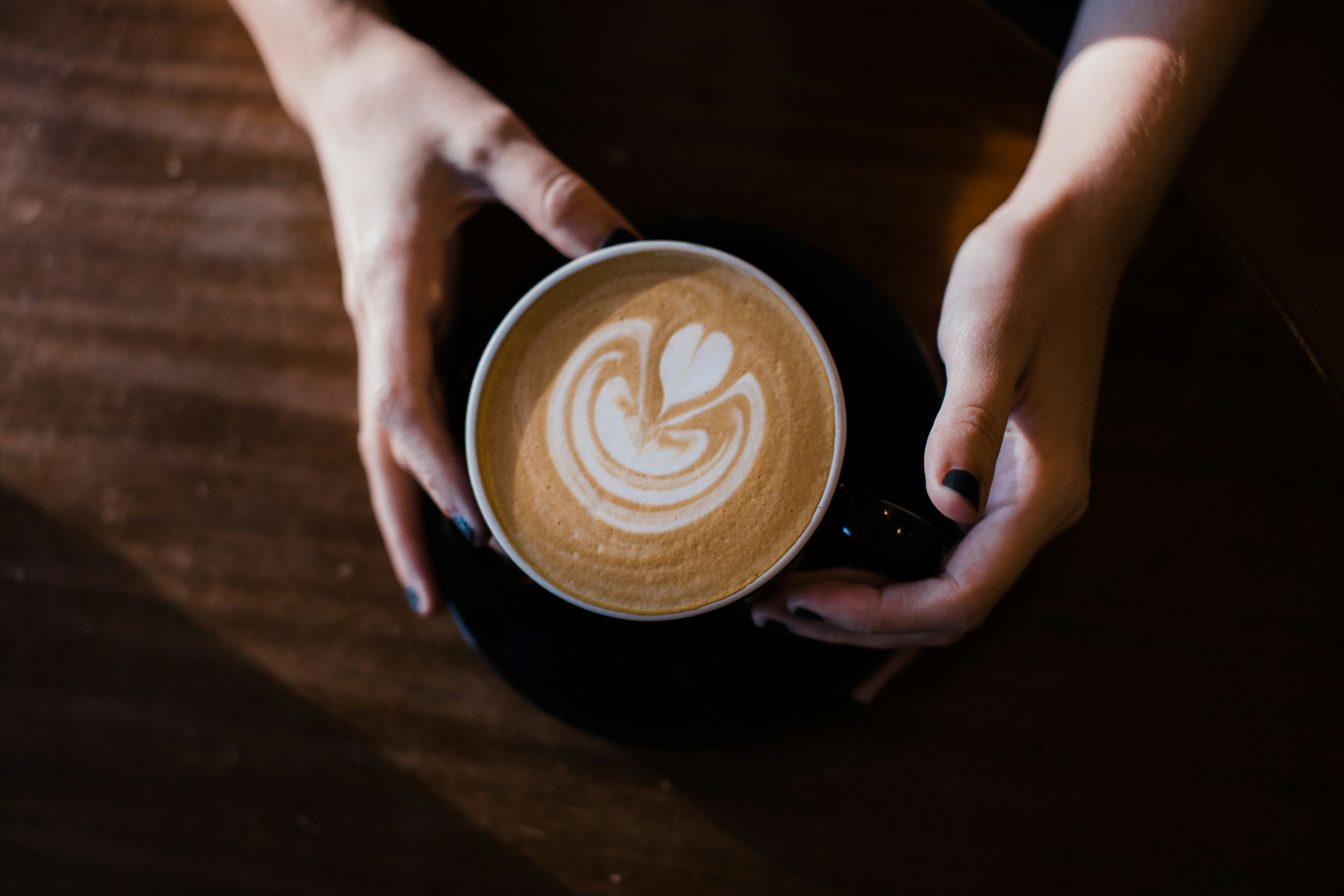 Hands gently holding a cup of latte with intricate foam art on a wooden table.