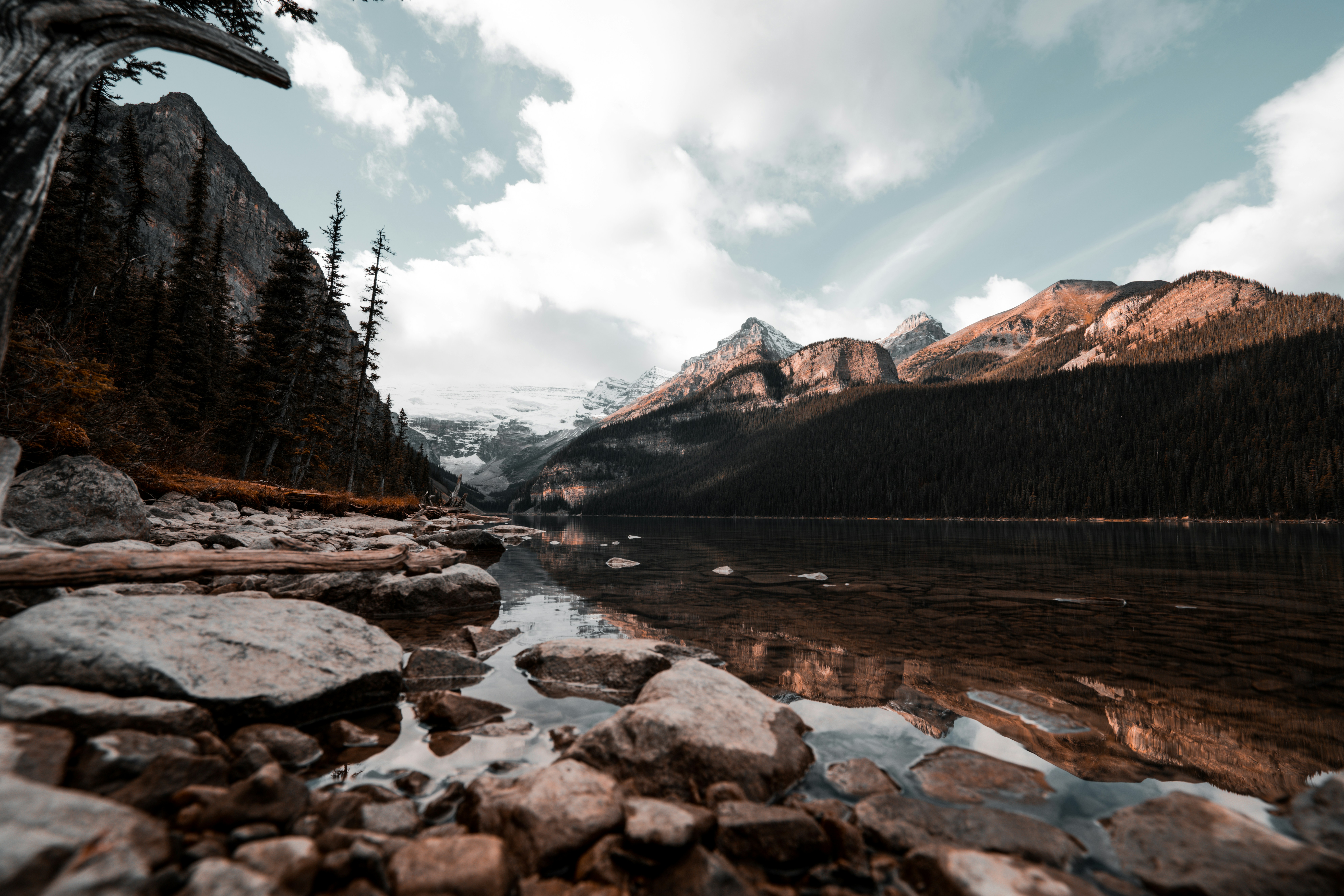 Tranquil lake reflecting majestic mountains under a partly cloudy sky, with rocky shoreline in the foreground.