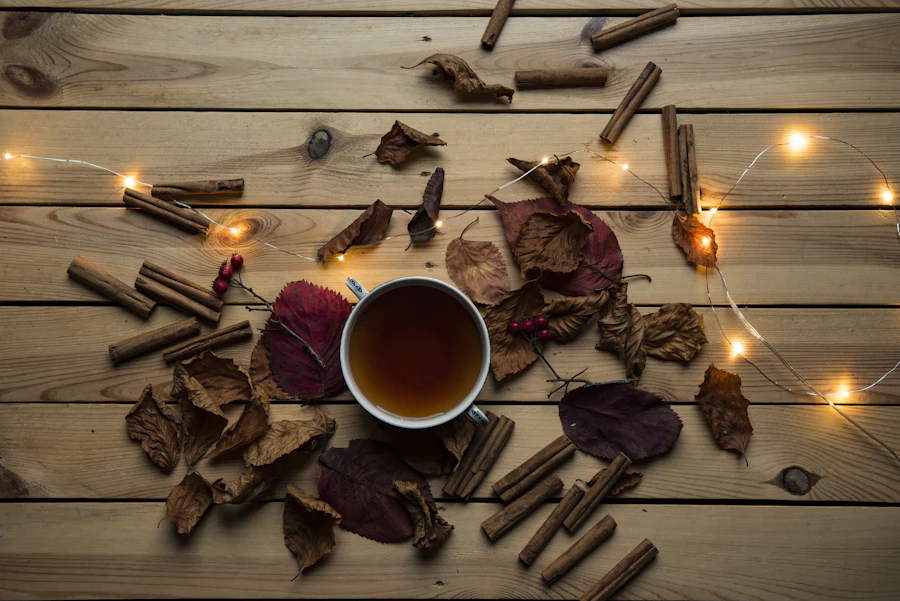 A cup of tea sits on a wooden plank table surrounded by dried autumn leaves, cinnamon sticks, and warm string lights.