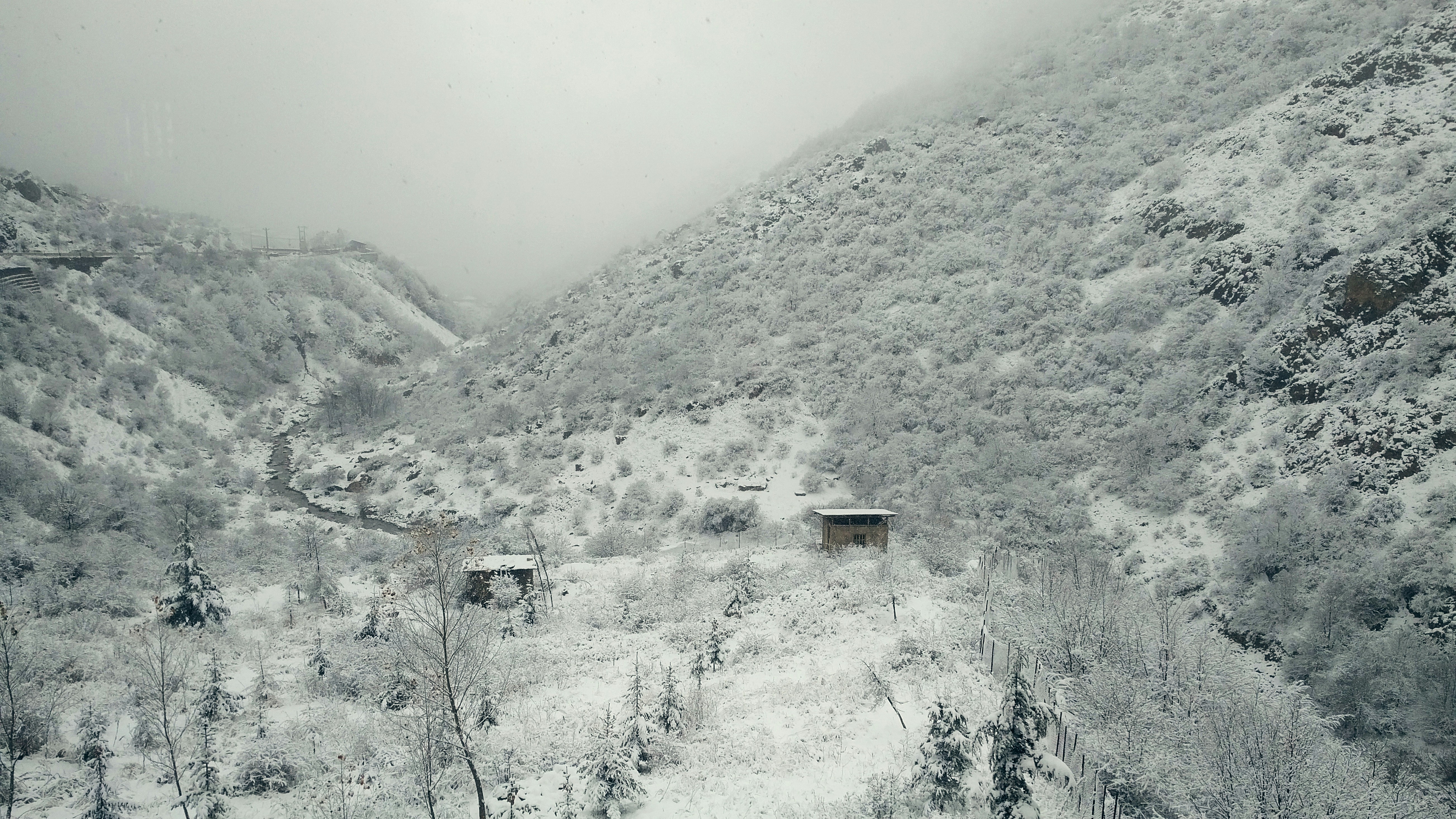 Snow-covered hills enveloping two rustic cabins, with a winding path disappearing into the misty distance. The tranquil scene evokes a sense of solitude and peace.