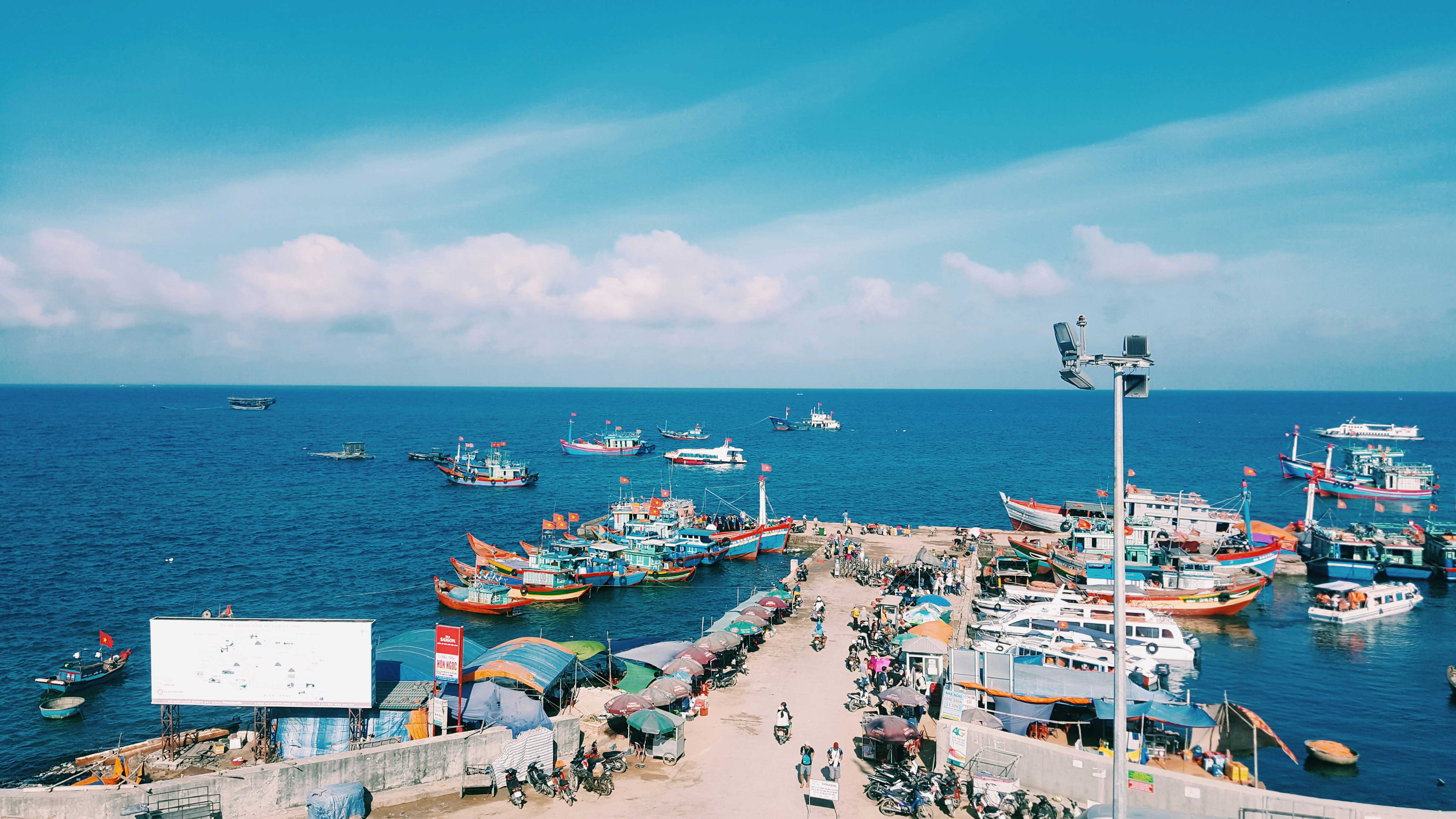 Photograph of a busy harbor with a wooden pier lined by colorful boats and market stalls extending into the blue sea. The expansive horizon and bright sky emphasize the scene's lively maritime activity.